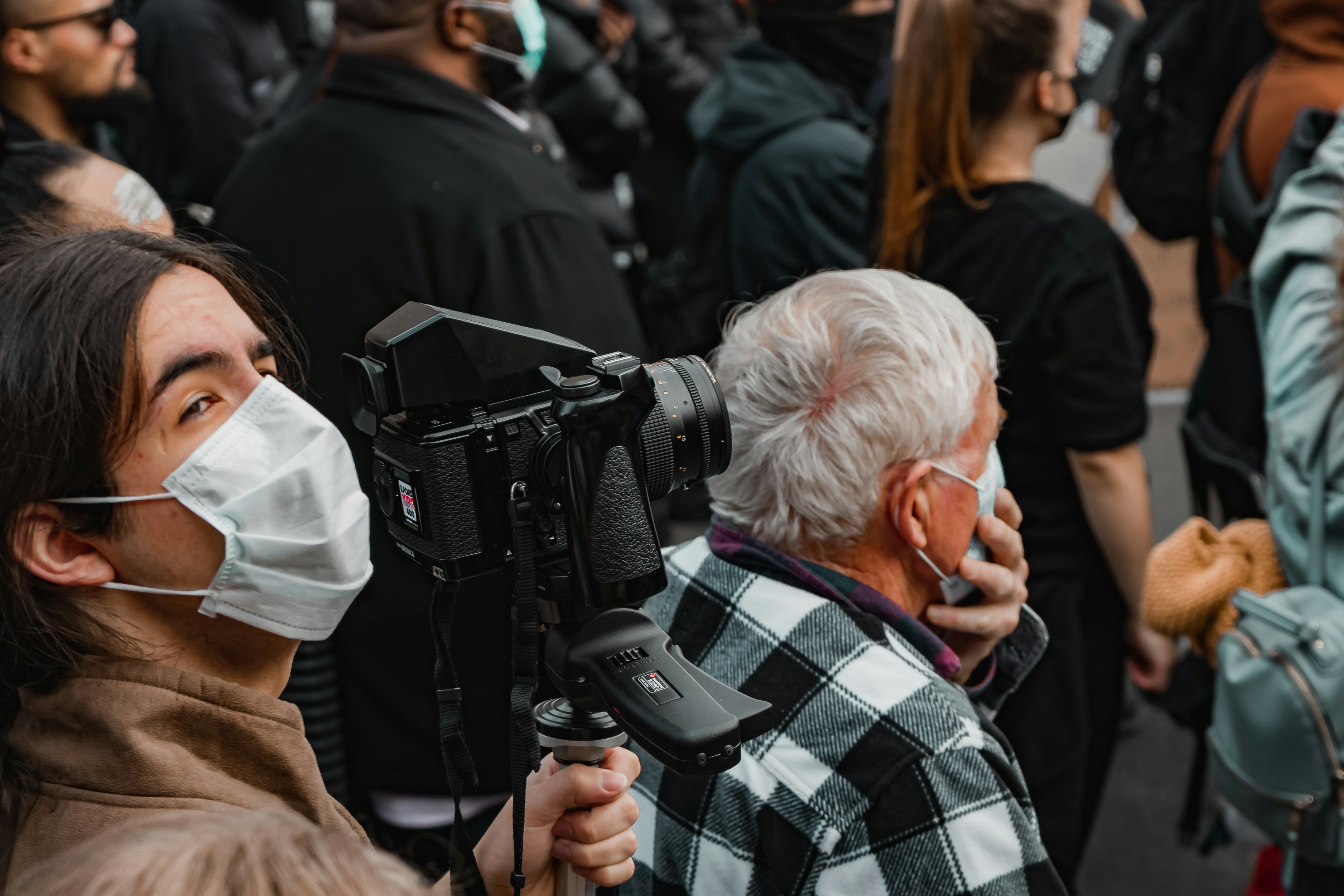 A young man with a camera smiles while wearing a mask, surrounded by a diverse crowd. The scene reflects a moment of connection amidst a gathering.