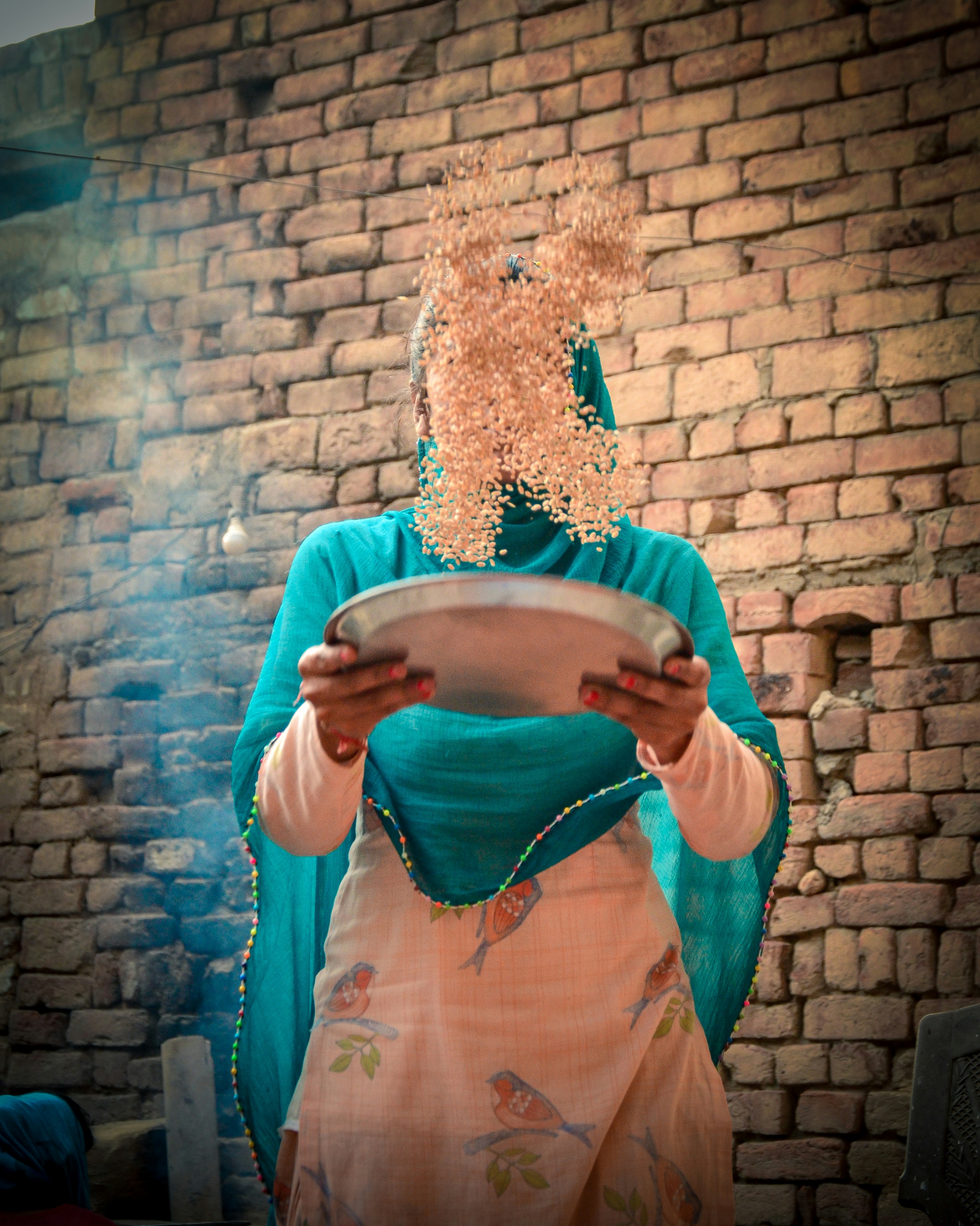 Woman in a vibrant teal shawl skillfully tossing grains in a rustic setting, showcasing a cultural culinary practice.