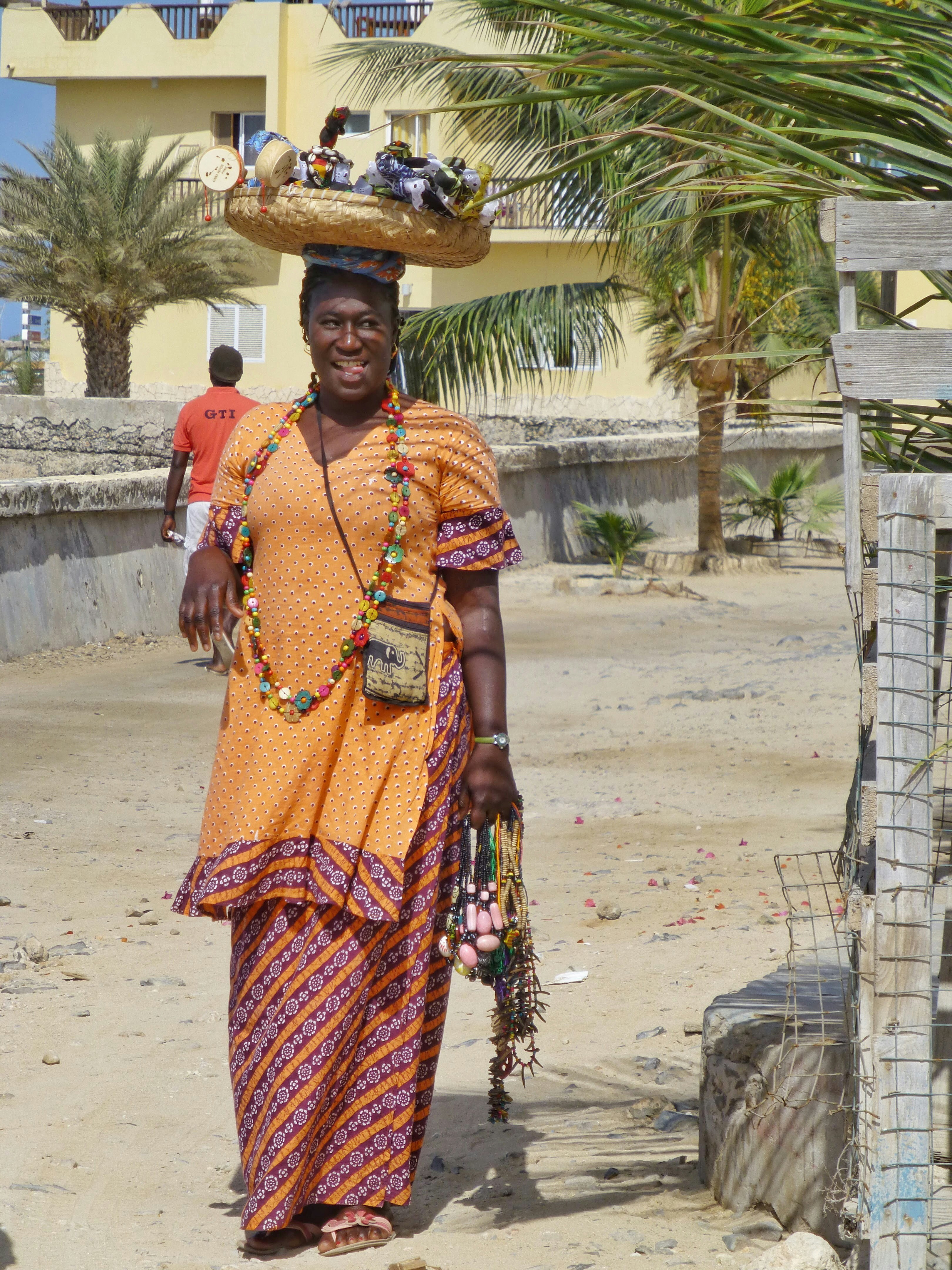 Woman in colorful traditional attire walking along a sandy path, carrying a basket on her head and adorned with jewelry. The backdrop features palm trees and a sunny coastal setting.