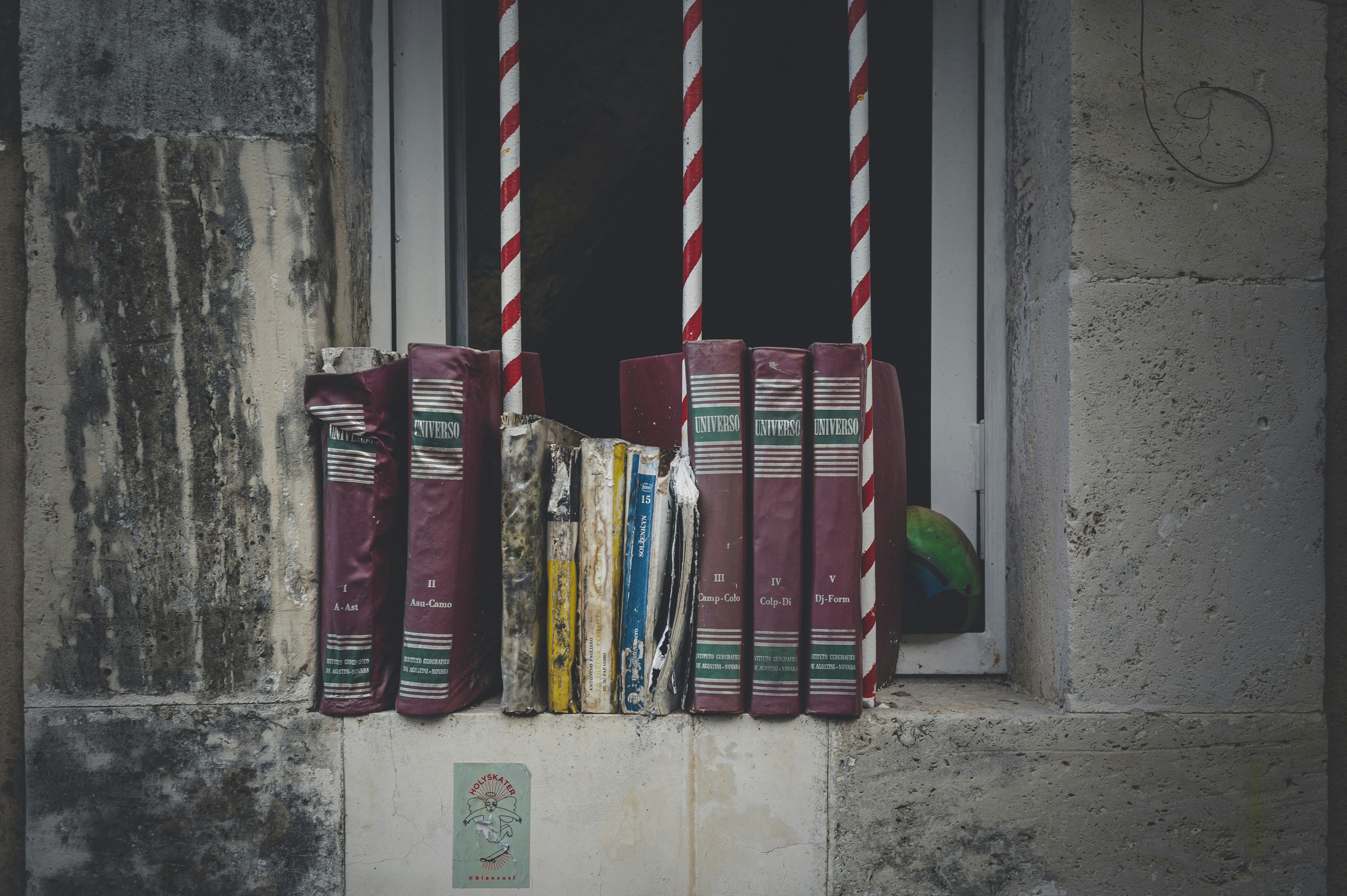 Weathered books resting on a window ledge of an aged stone building, with red and white striped bars.