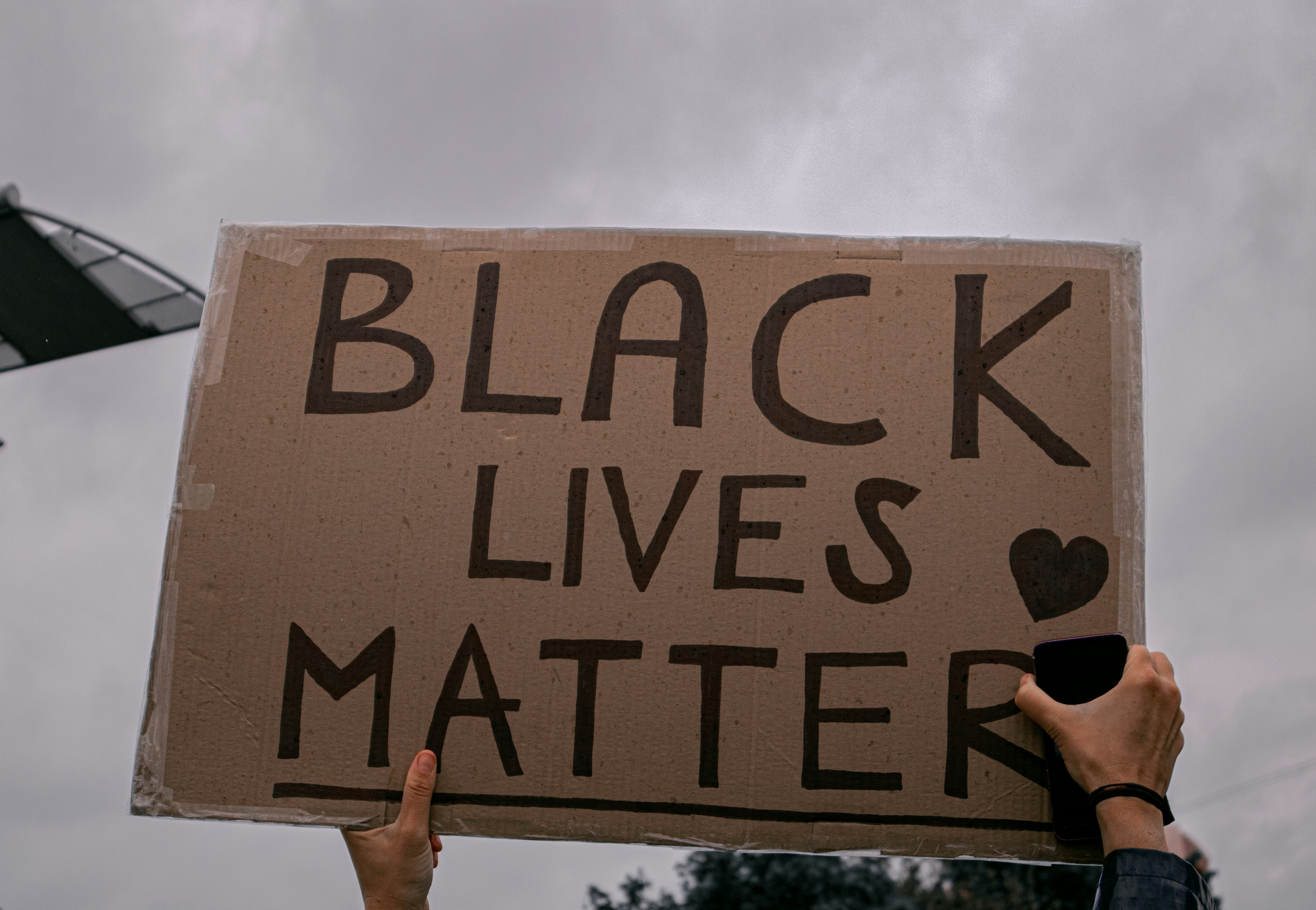 Hands holding a cardboard sign displaying the message 'BLACK LIVES MATTER' against a cloudy backdrop.