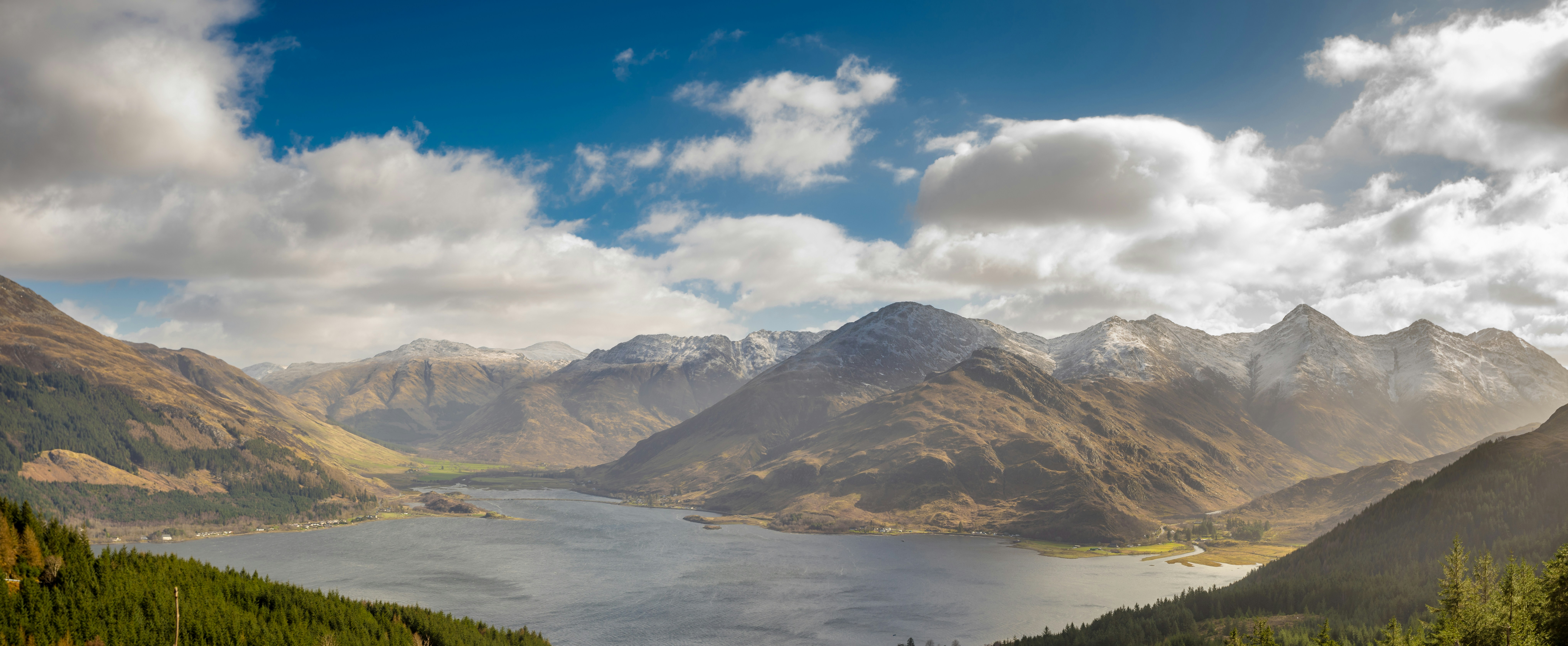 Expansive mountain range with a serene lake below, under a sky dotted with fluffy clouds.