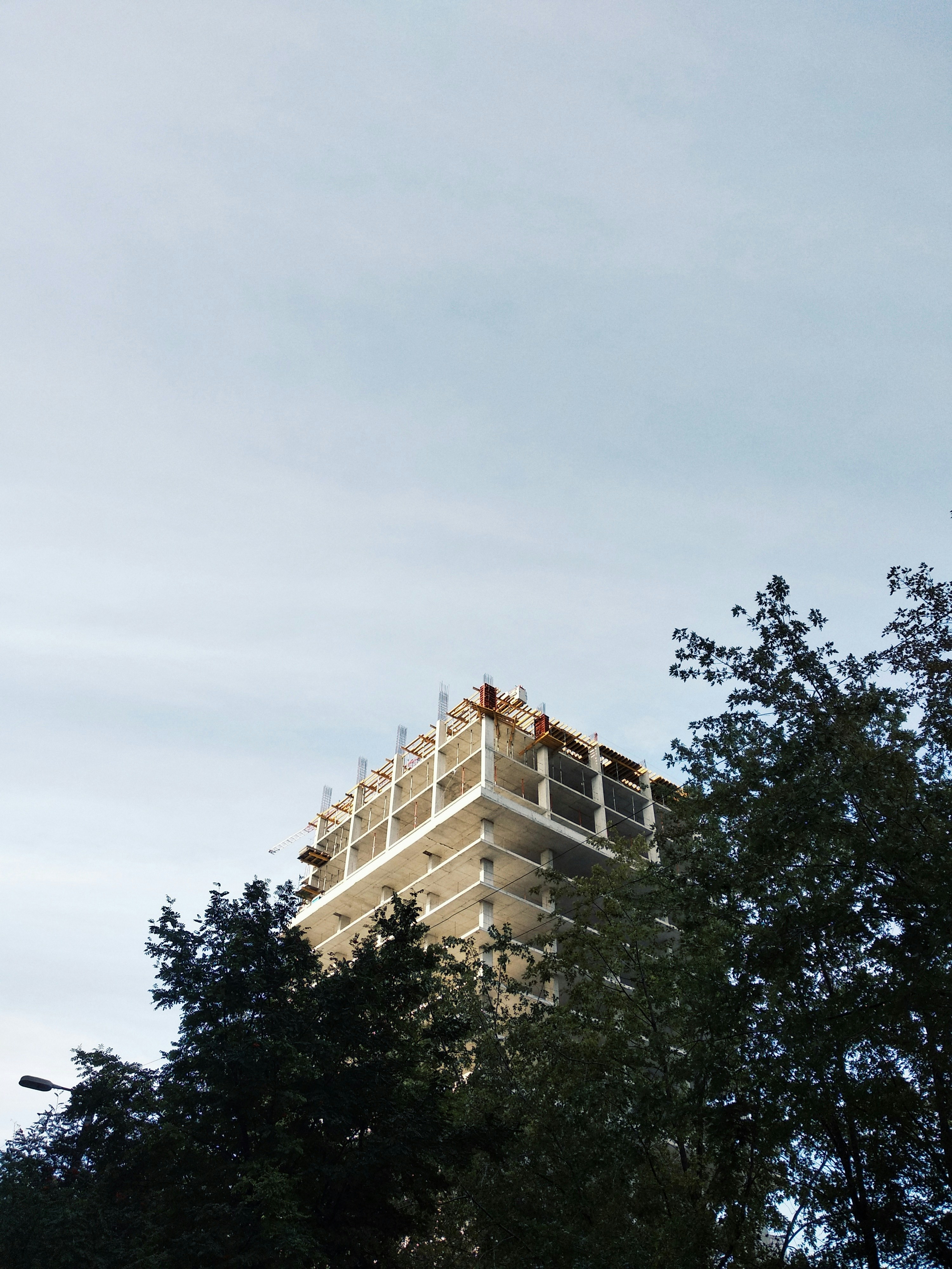 Concrete building under construction, framed by surrounding trees, reaching toward a cloudy sky.