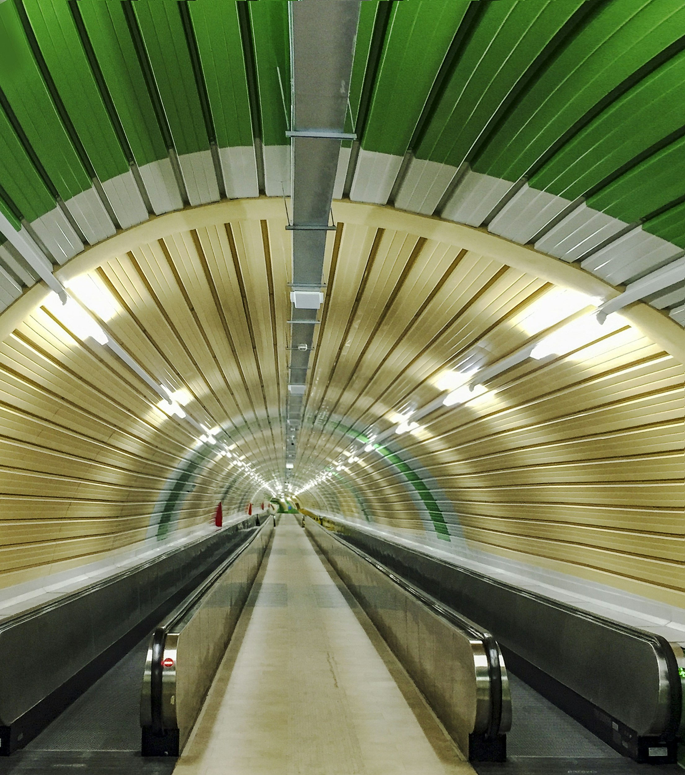 Futuristic escalator corridor featuring dynamic green and beige curves, leading into the distance. Bright lighting enhances the architectural design.