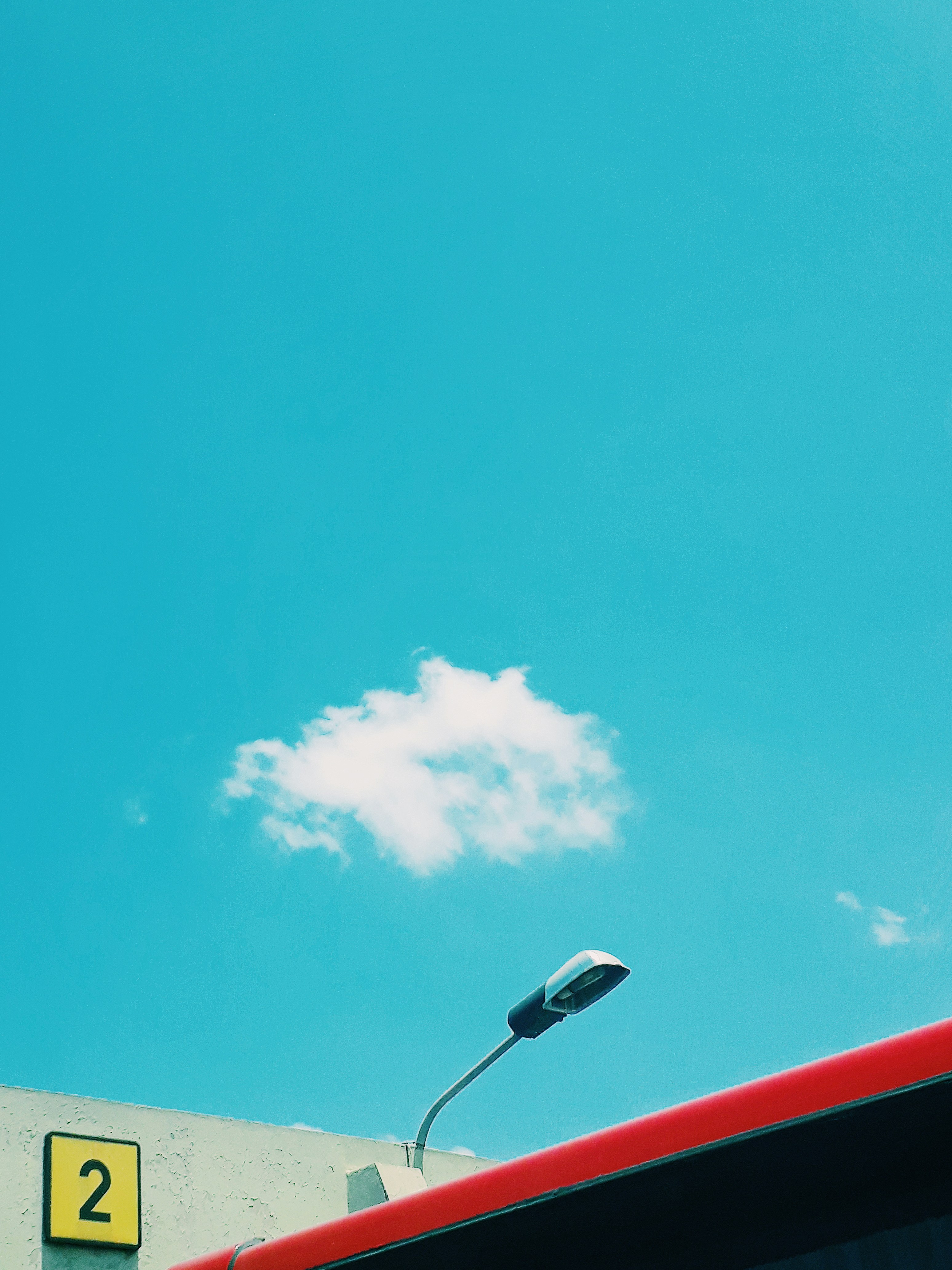 Minimalist photograph of a bright blue sky with a curved streetlamp over a diagonal red edge and a small white cloud.