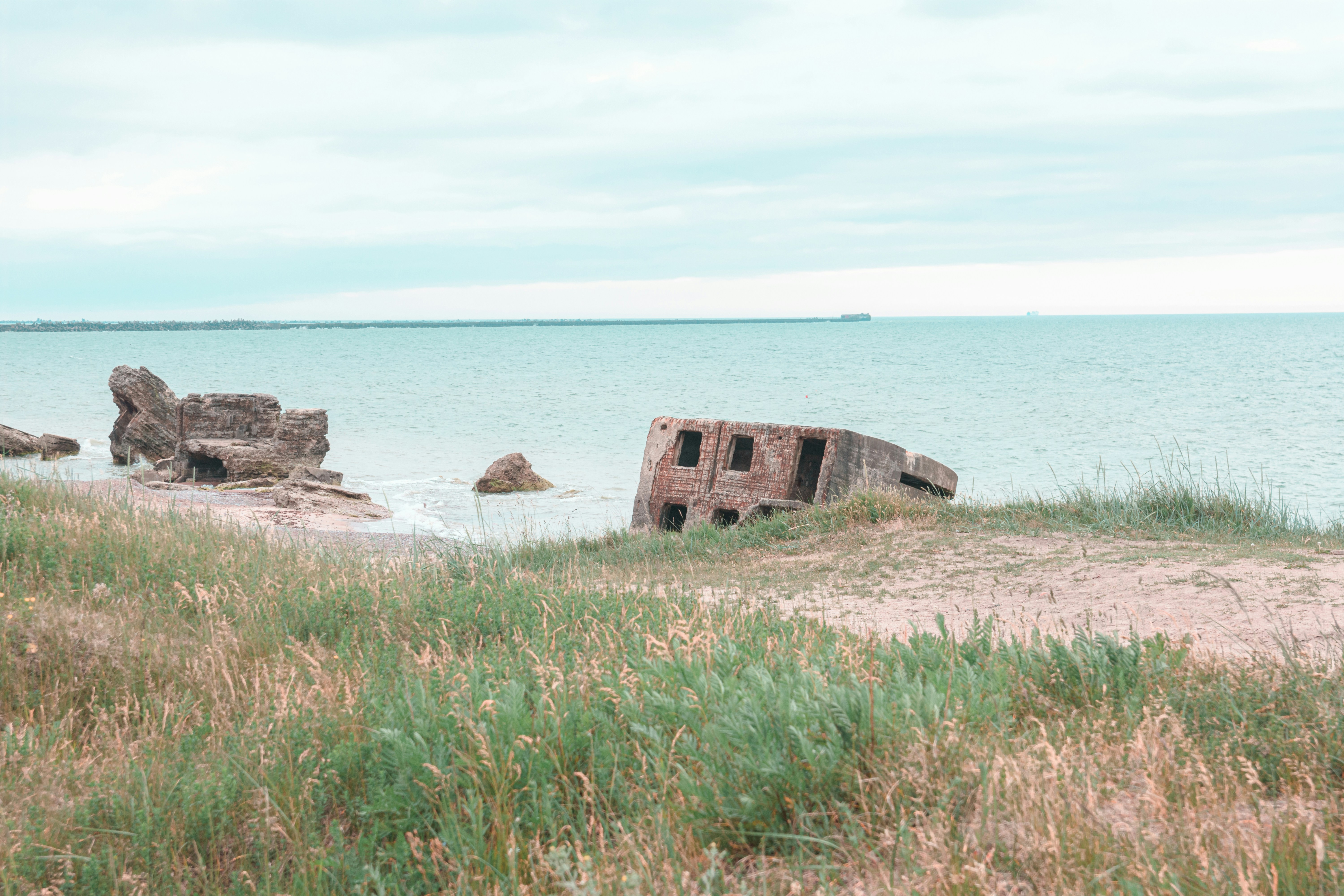 An abandoned shipwreck partially submerged near a tranquil shoreline, surrounded by grass and rocky formations. The calm sea reflects a soft, muted sky.