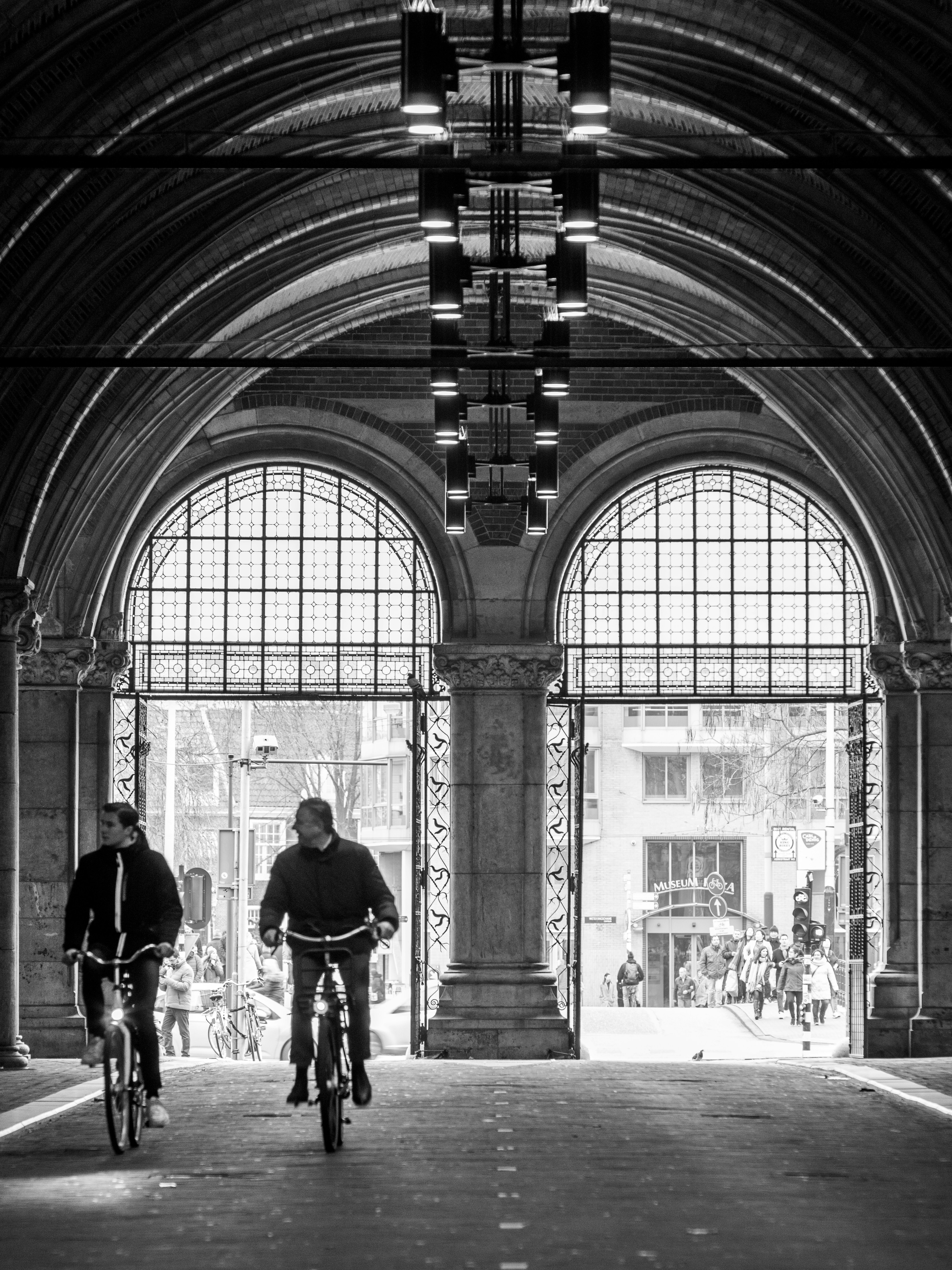 Two cyclists navigate a grand archway adorned with intricate details, framed by large, arched windows. The scene captures a blend of movement and stillness in a bustling urban environment.