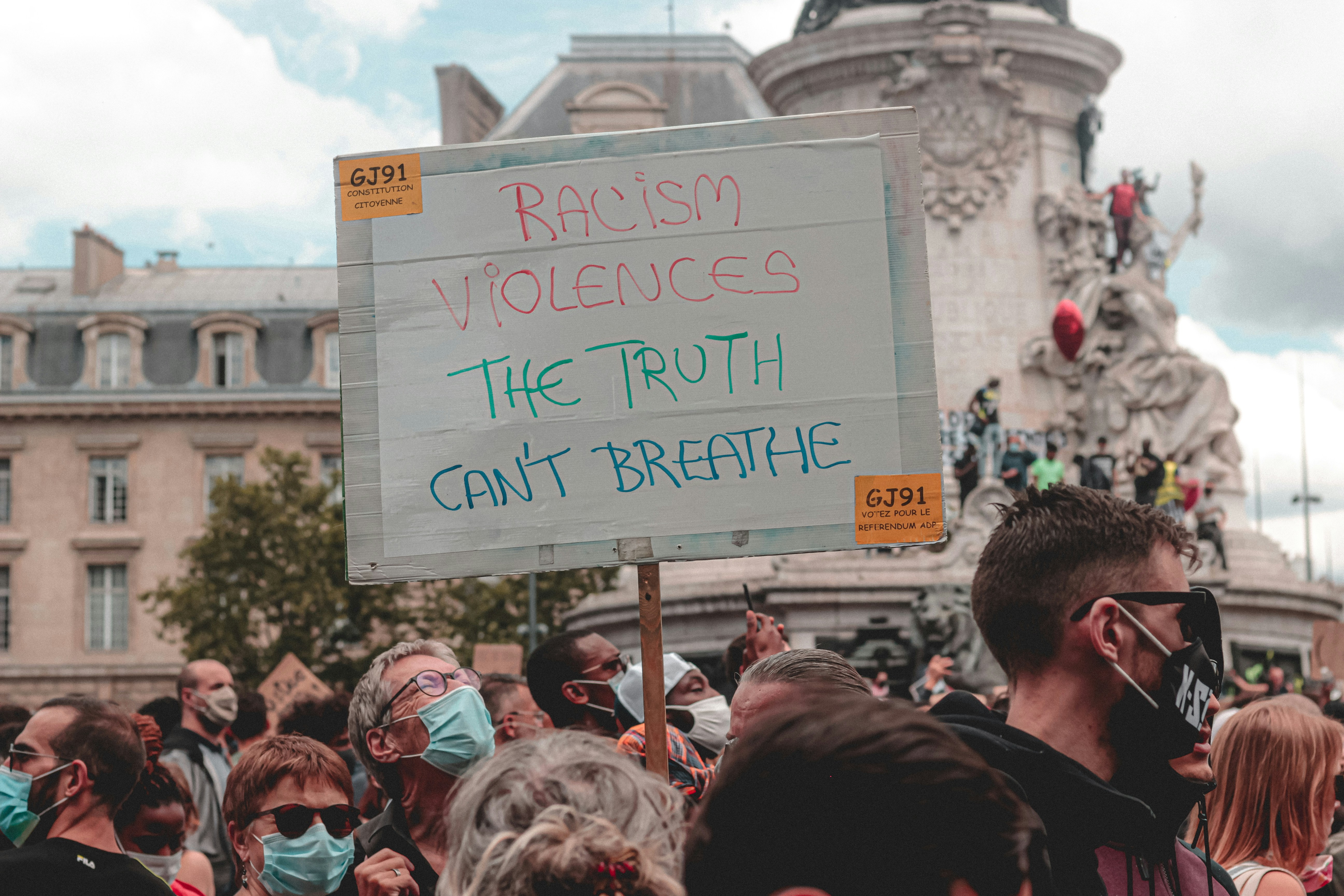 Protesters holding a sign demanding justice against racism and violence, set against a historic backdrop with a diverse crowd wearing masks.