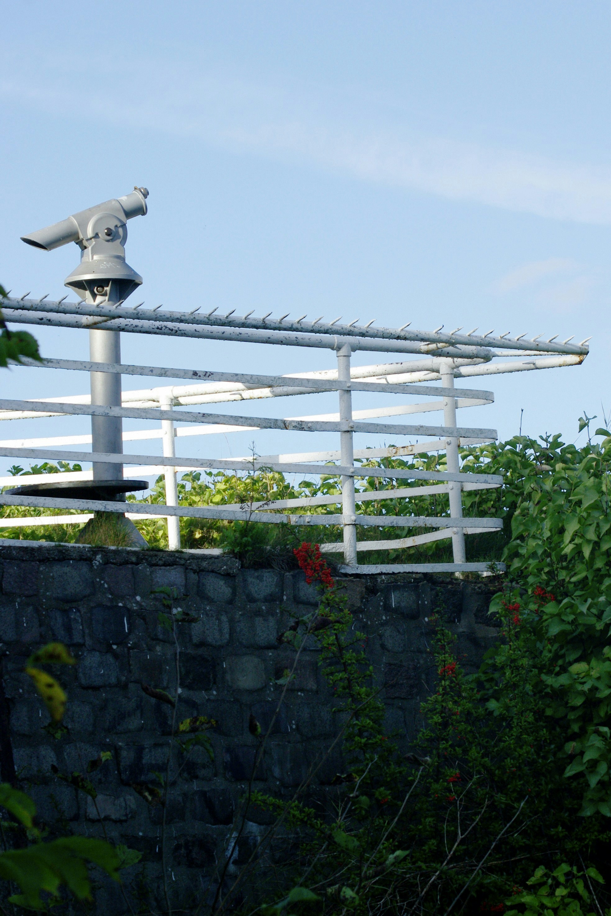 A weathered metal telescope-like device sits atop a railing above a stone wall, with greenery and a clear blue sky in the background.