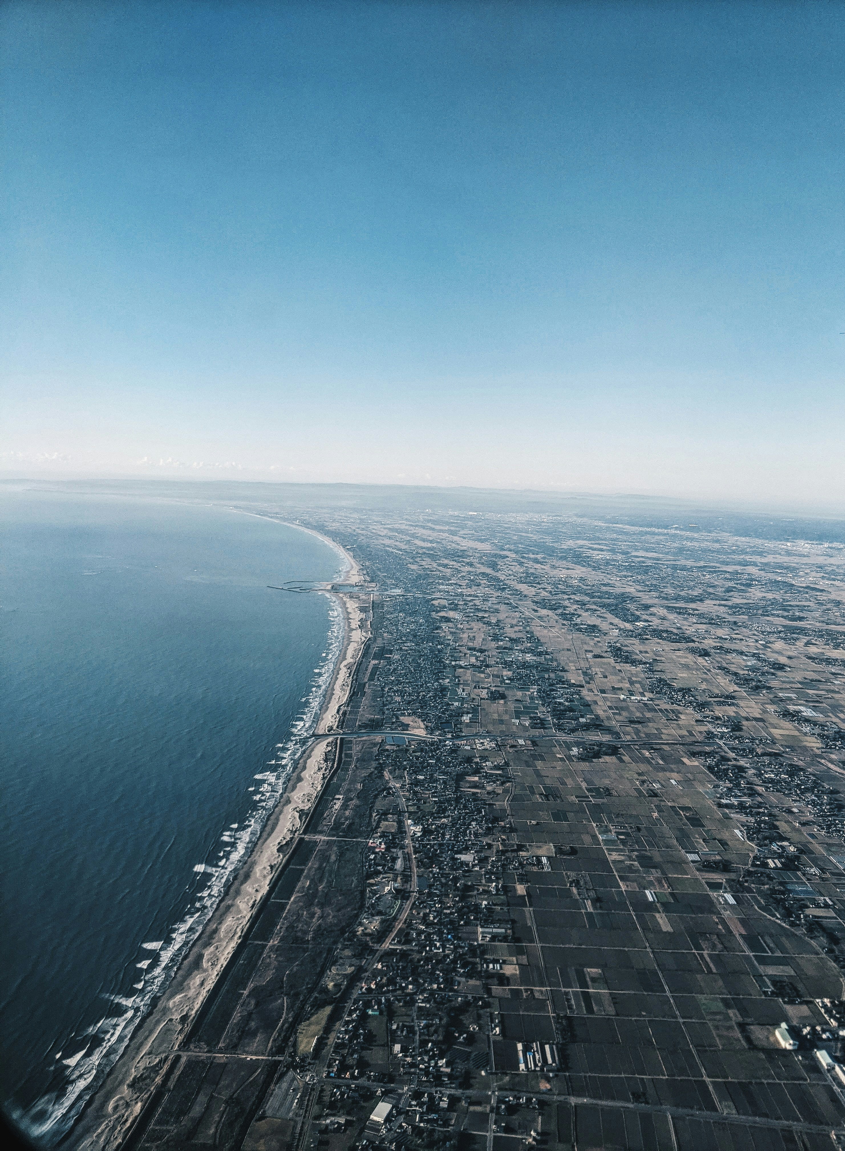 Aerial view showcasing the dynamic coastline meeting the expansive ocean, with agricultural fields and urban areas visible in the distance.