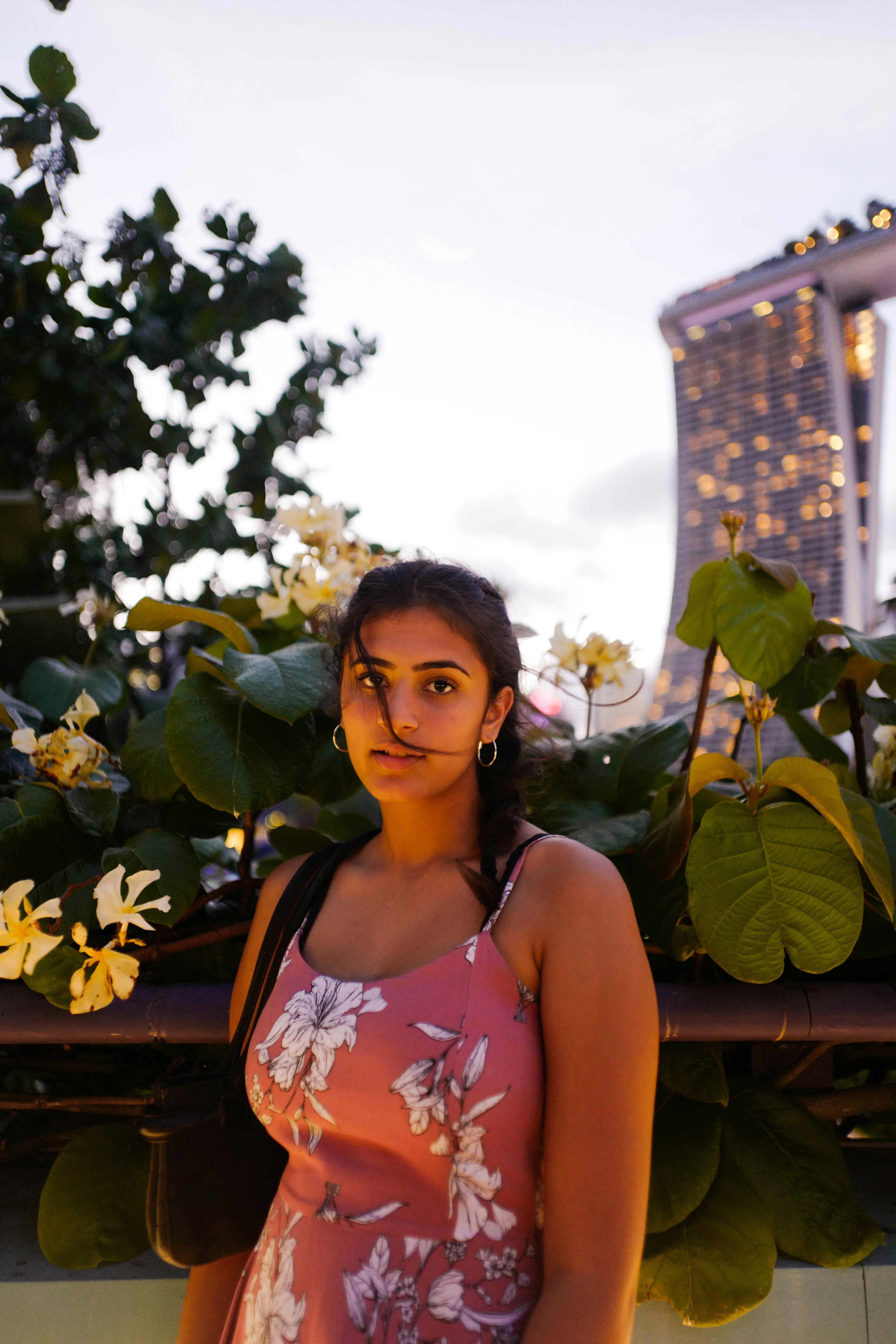 Young woman with a floral dress stands amidst vibrant foliage and soft evening lights, evoking a serene atmosphere.