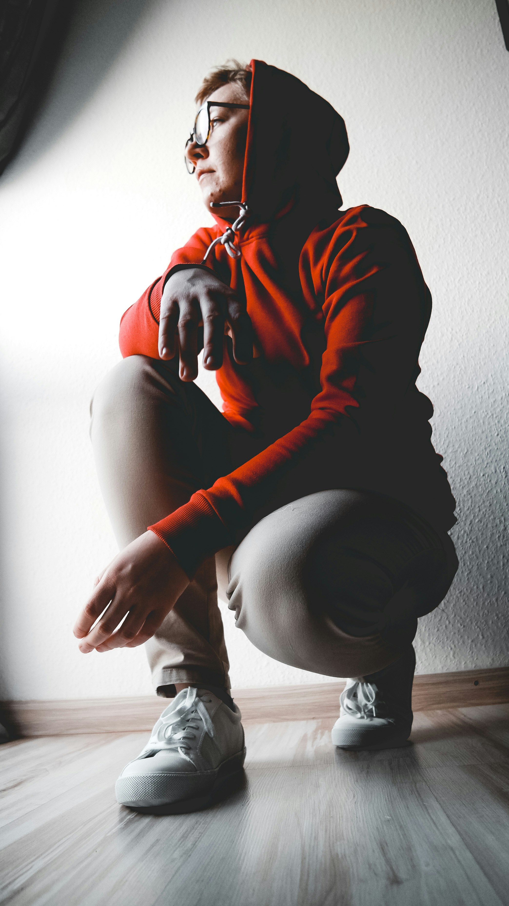 Low-angle portrait in a red hoodie: a pensive crouch against a sunlit wall