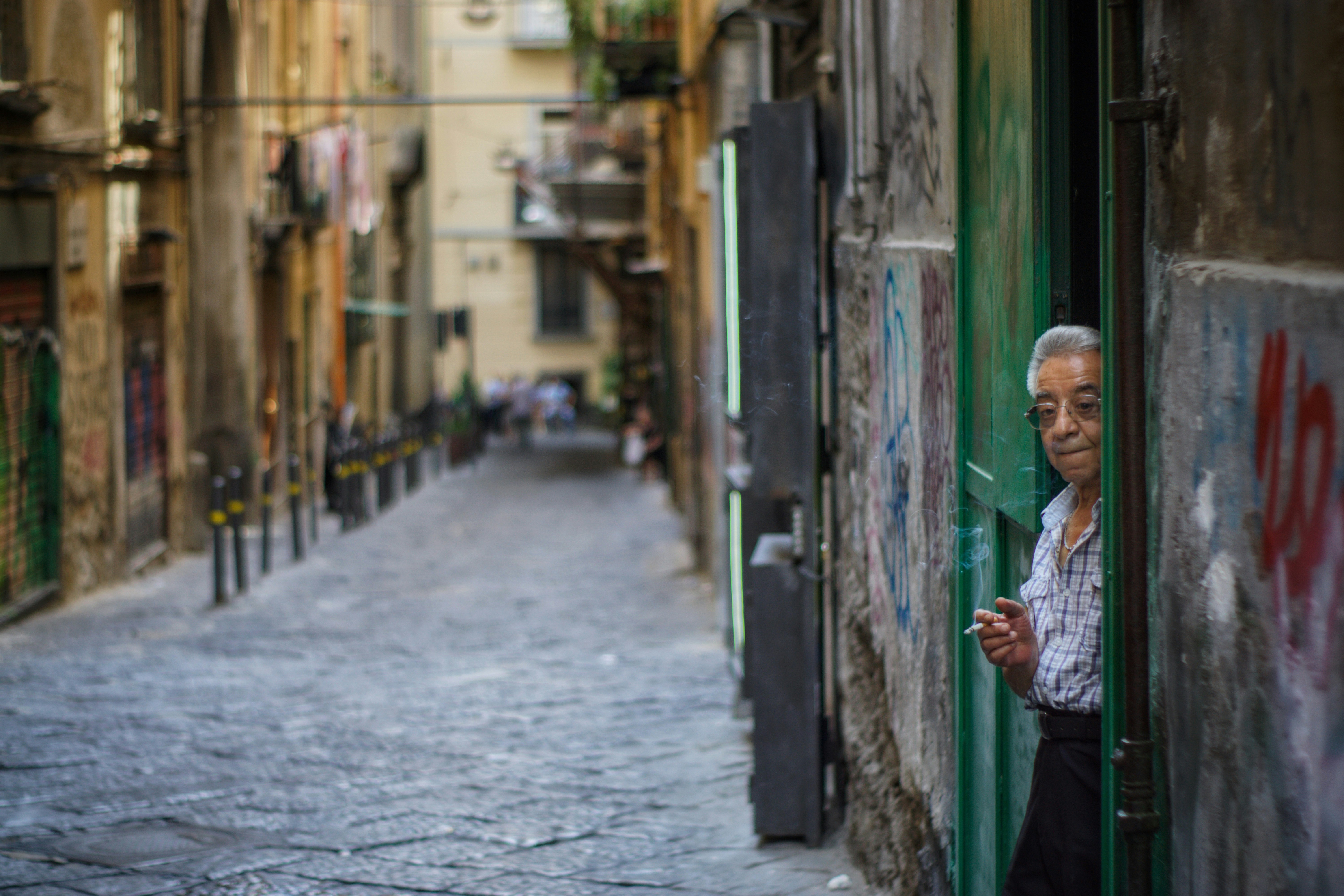 Doorway Observer: An elderly man peeks from a green doorway on a sunlit cobblestone street