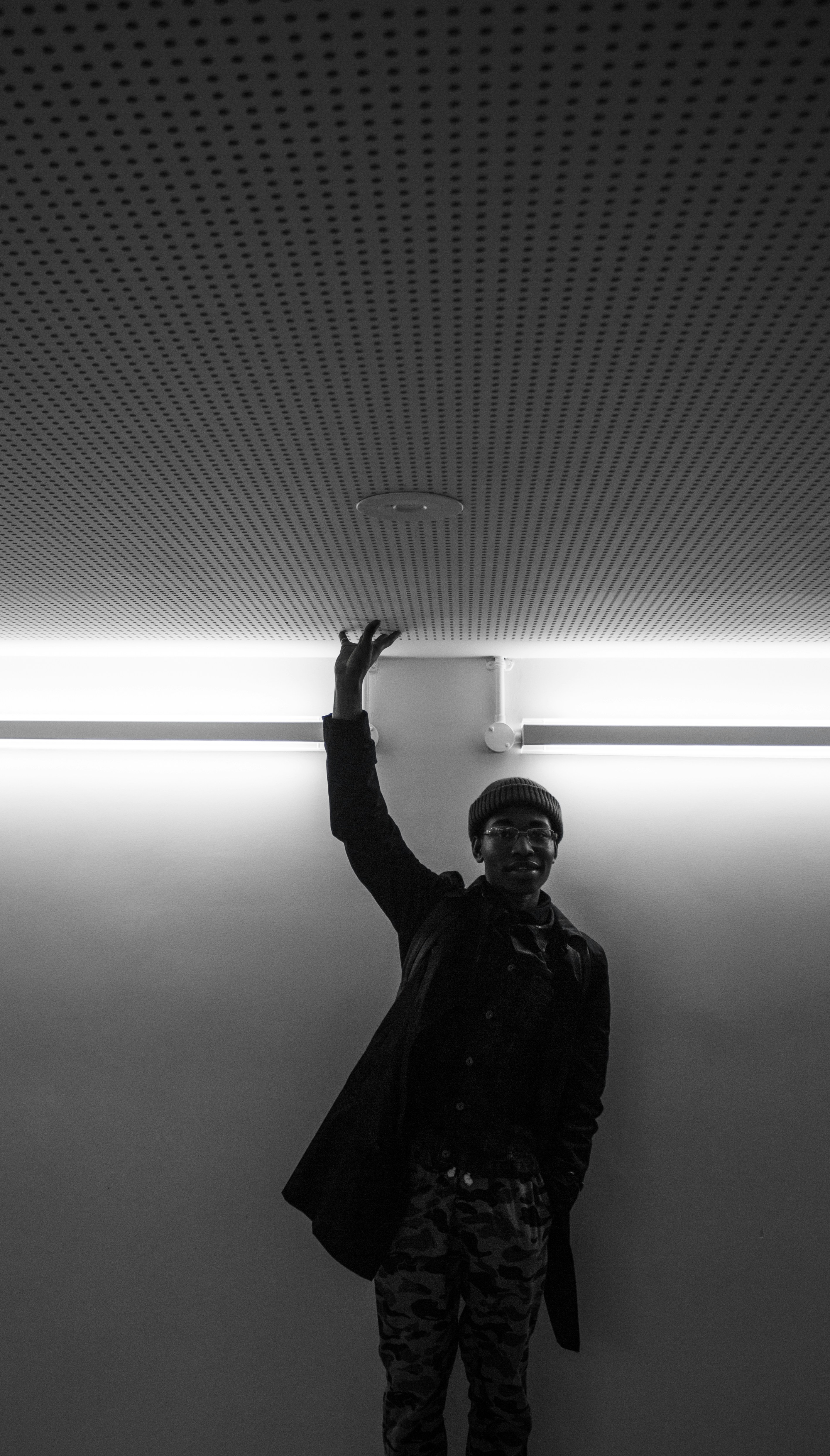 Monochrome Corridor Reach: A Lone Figure Touches the Perforated Ceiling