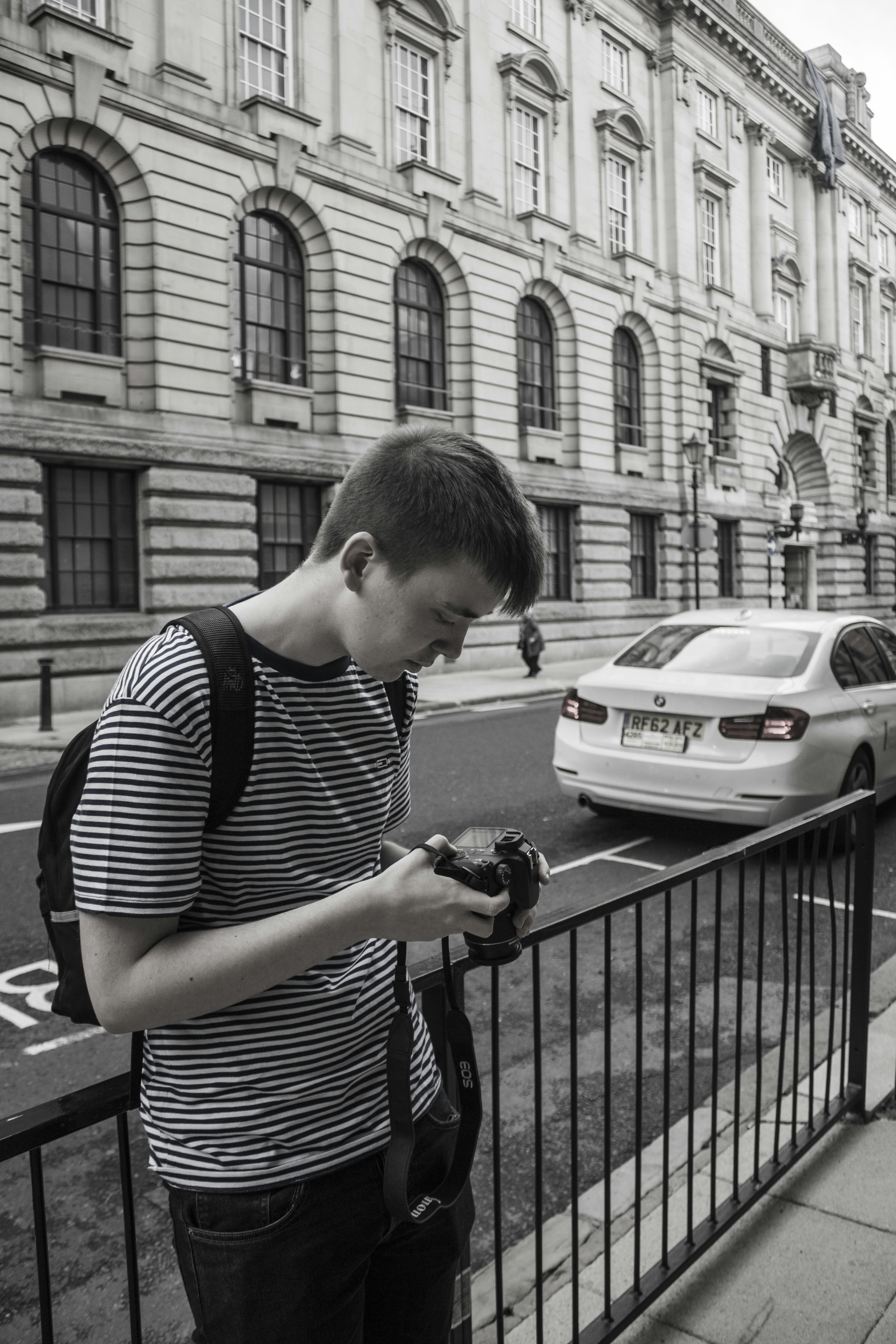 Young photographer adjusting camera settings on a city street, surrounded by historic architecture and modern vehicles.