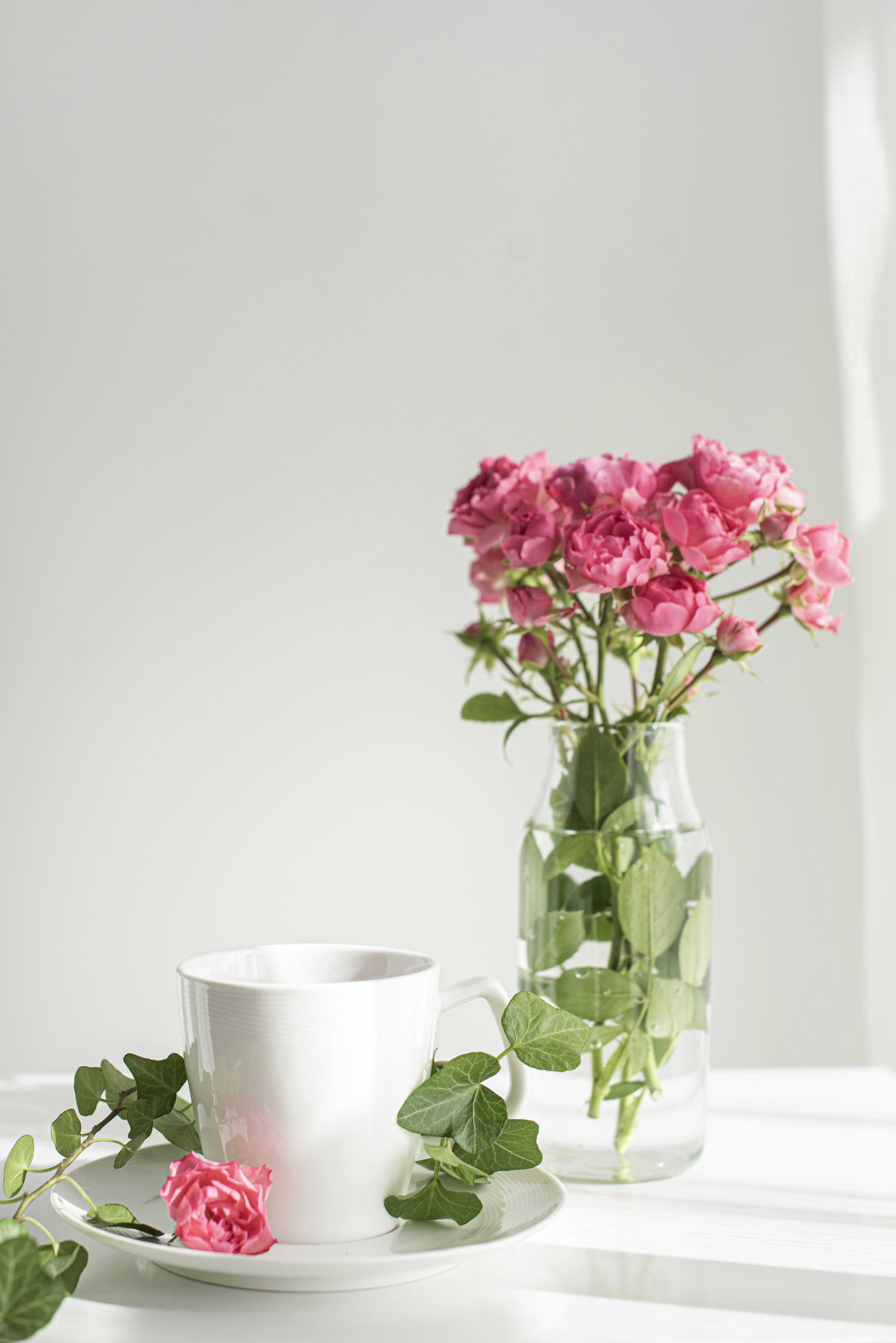 A white cup adorned with a pink rose sits beside a vase filled with blooming pink roses, capturing a tranquil tea moment. The soft light enhances the delicate colors and textures.