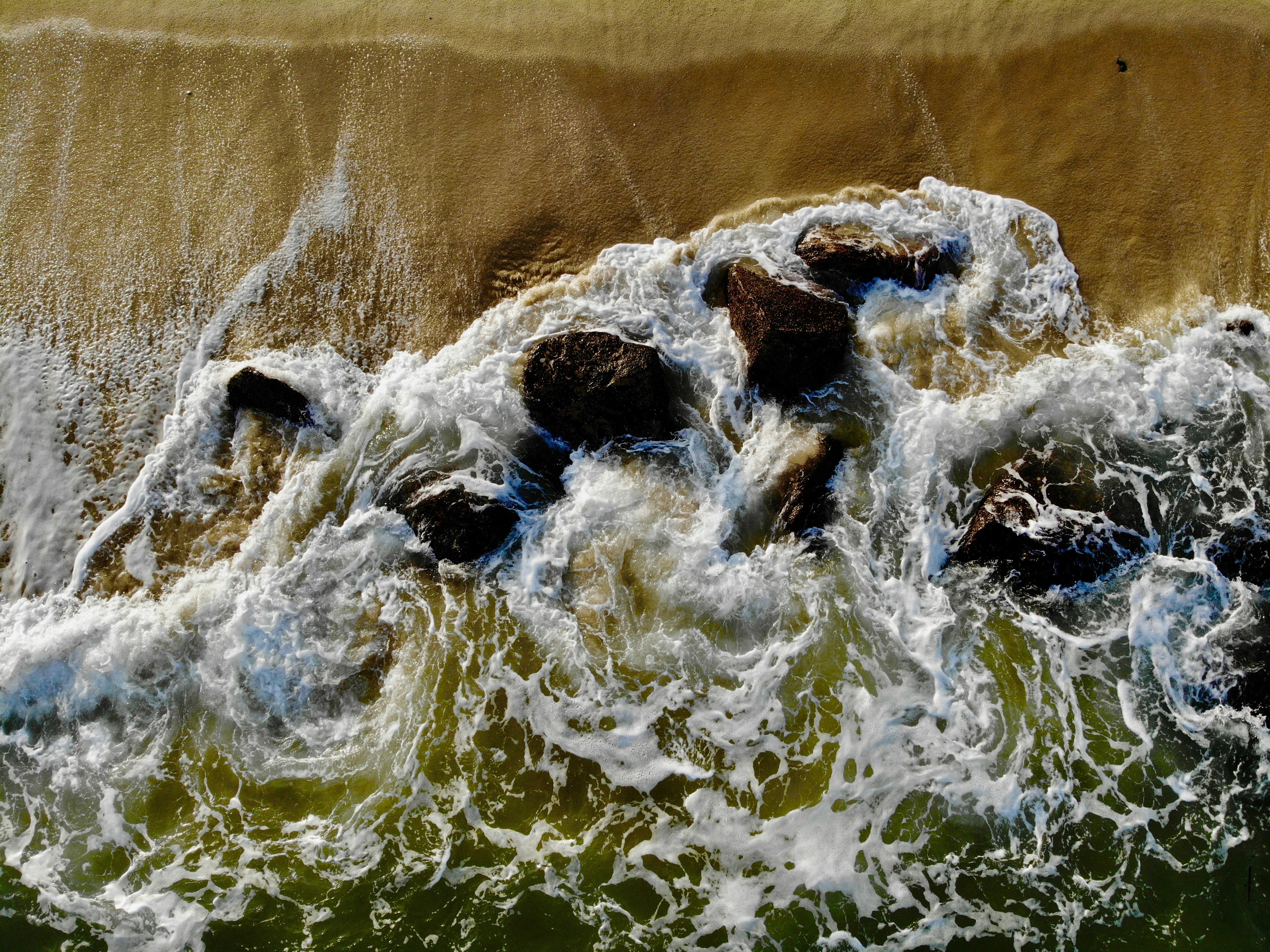 Aerial view of foamy waves crashing over rocks on a sandy beach.