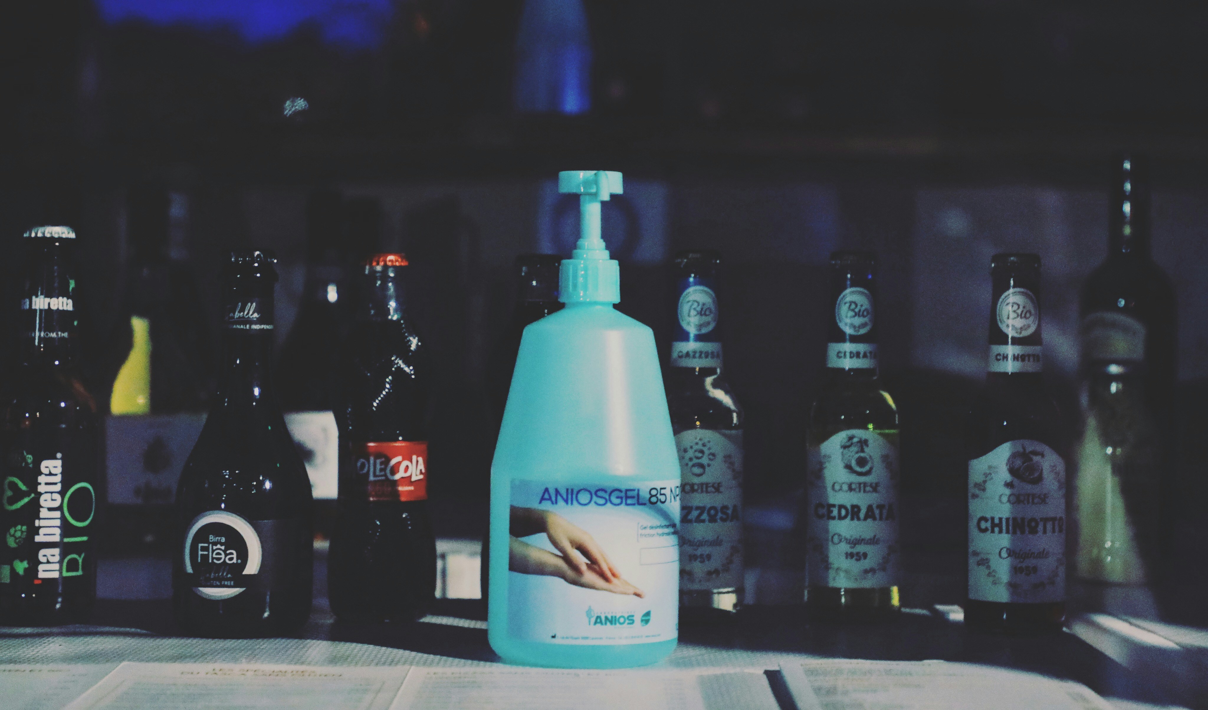 A prominent bottle of hand sanitizer stands out among a lineup of various alcoholic beverages on a bar countertop.
