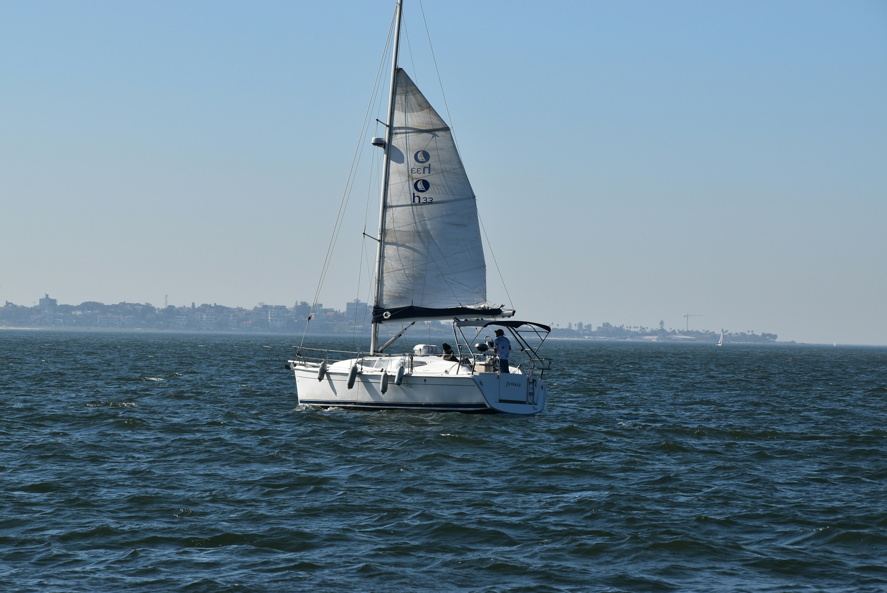 A sleek sailboat gliding across tranquil waters under a clear blue sky, with a distant city skyline faintly visible on the horizon.