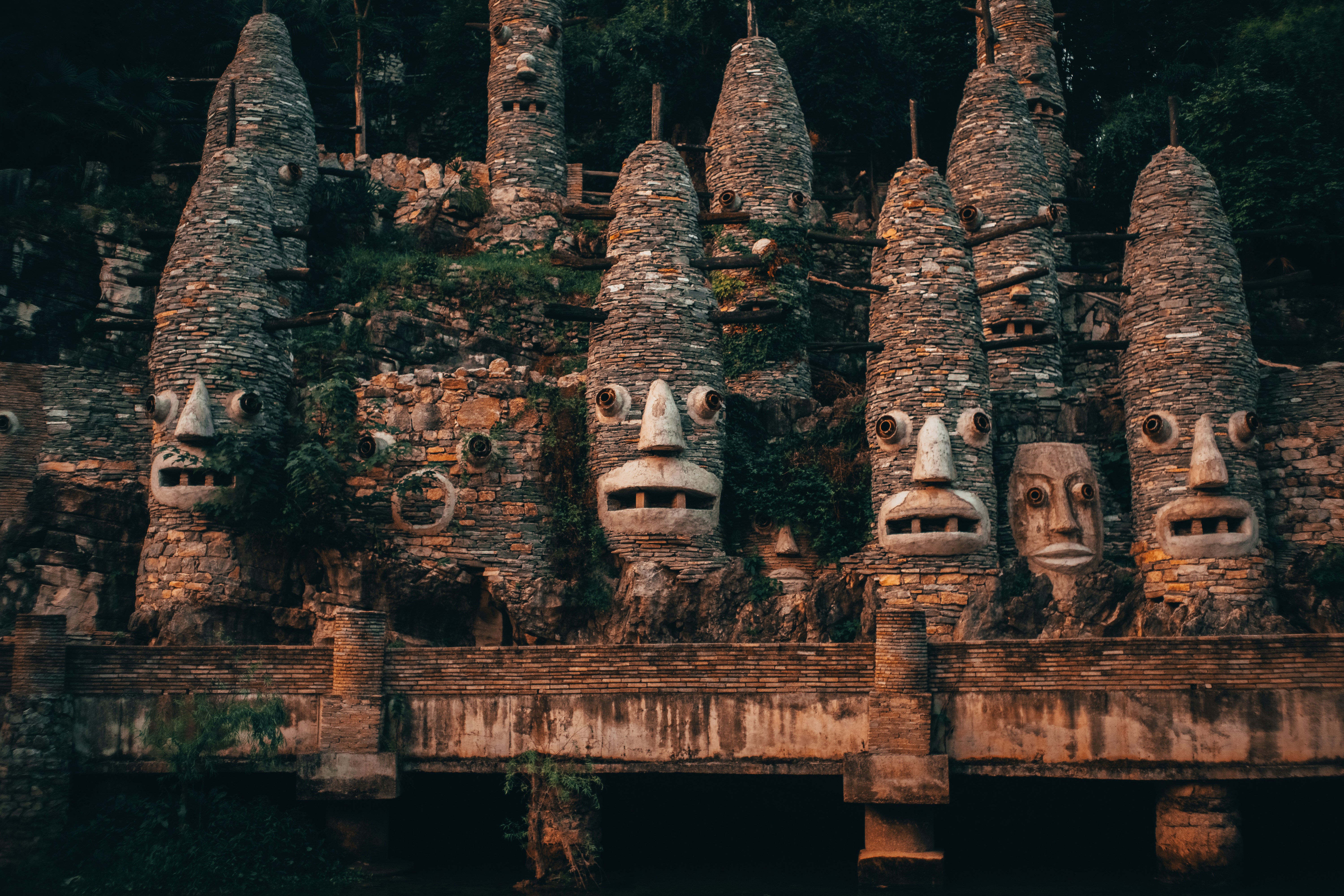 A series of intricately carved stone faces emerge from a moss-covered cliff, surrounded by lush greenery. Their expressions evoke a sense of ancient mystery.