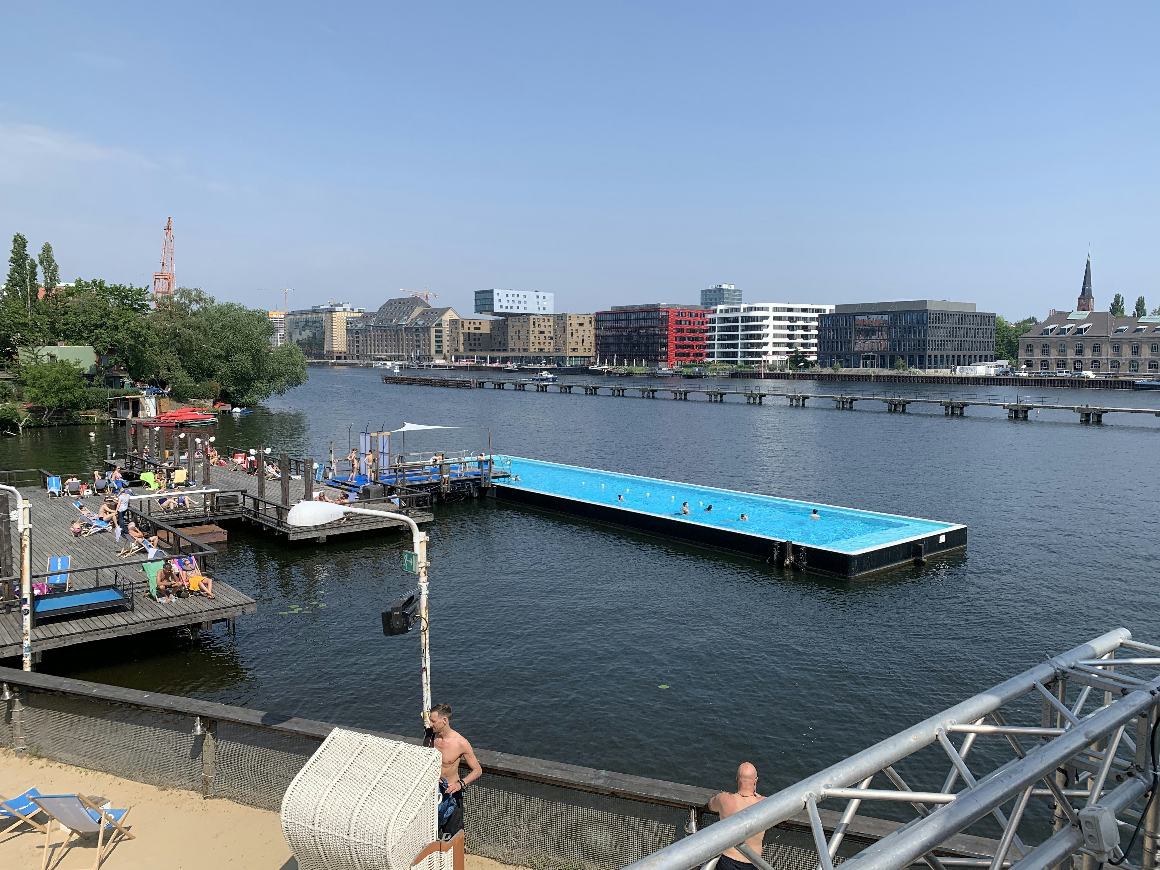 Vibrant swimming pool surrounded by modern architecture and lush greenery, showcasing a lively urban setting. The scene captures leisure activities along the water's edge.