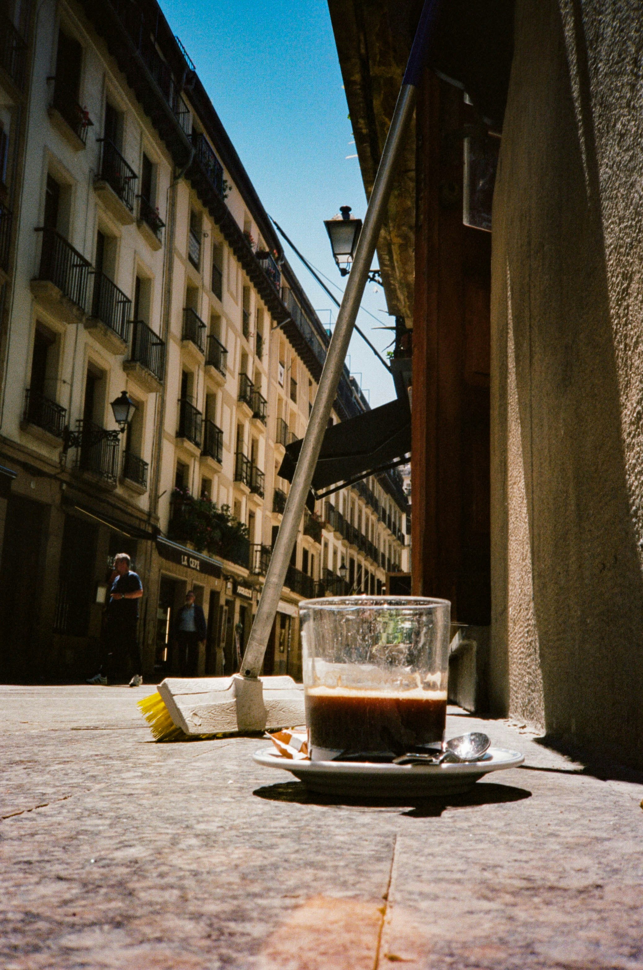 Half-drunk coffee resting on a plate beside a broom on a sunlit street, capturing the essence of everyday life in a bustling city.