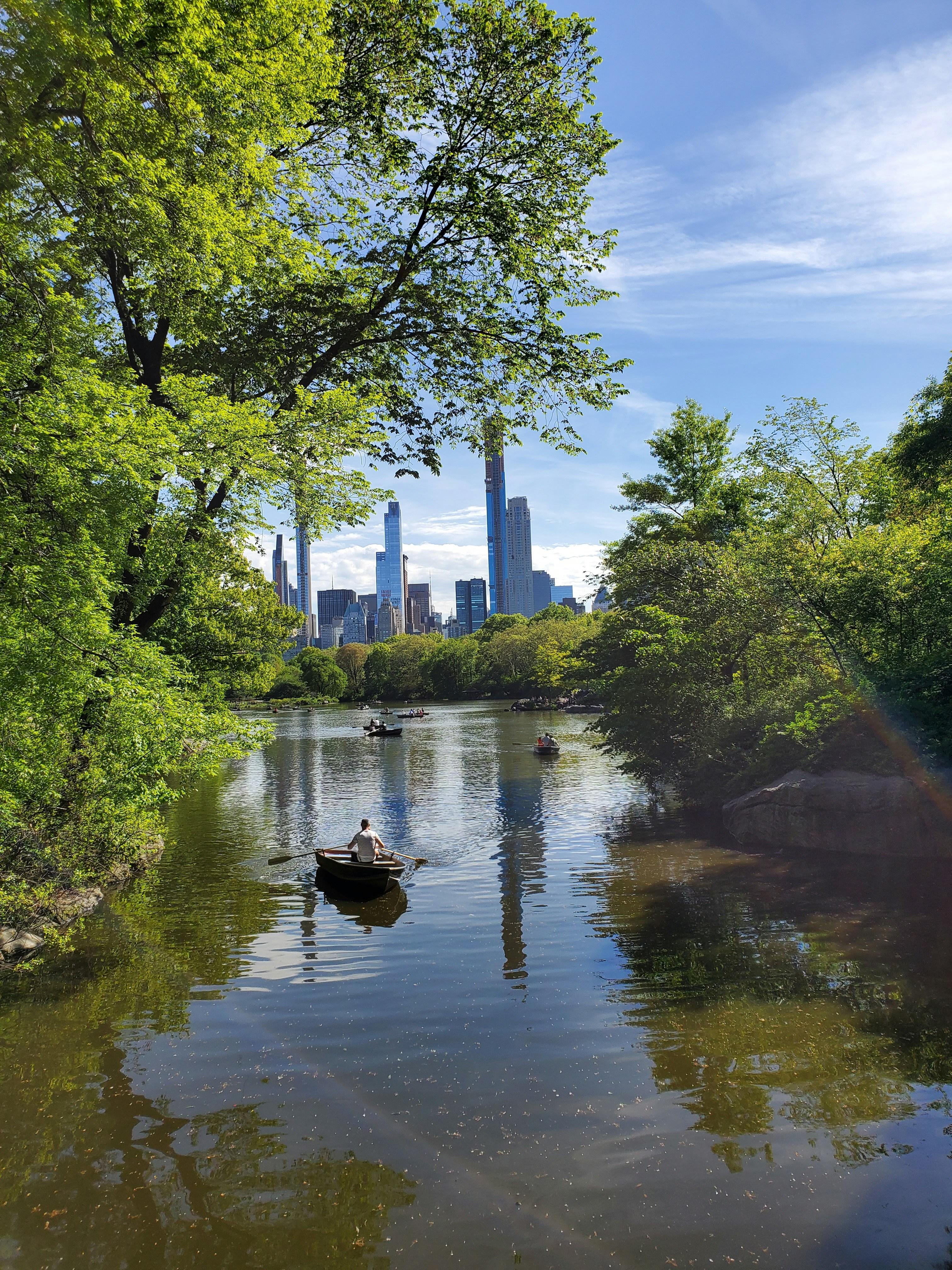 Rowboats glide peacefully on a reflective lake framed by lush greenery and towering city skyscrapers in the background.