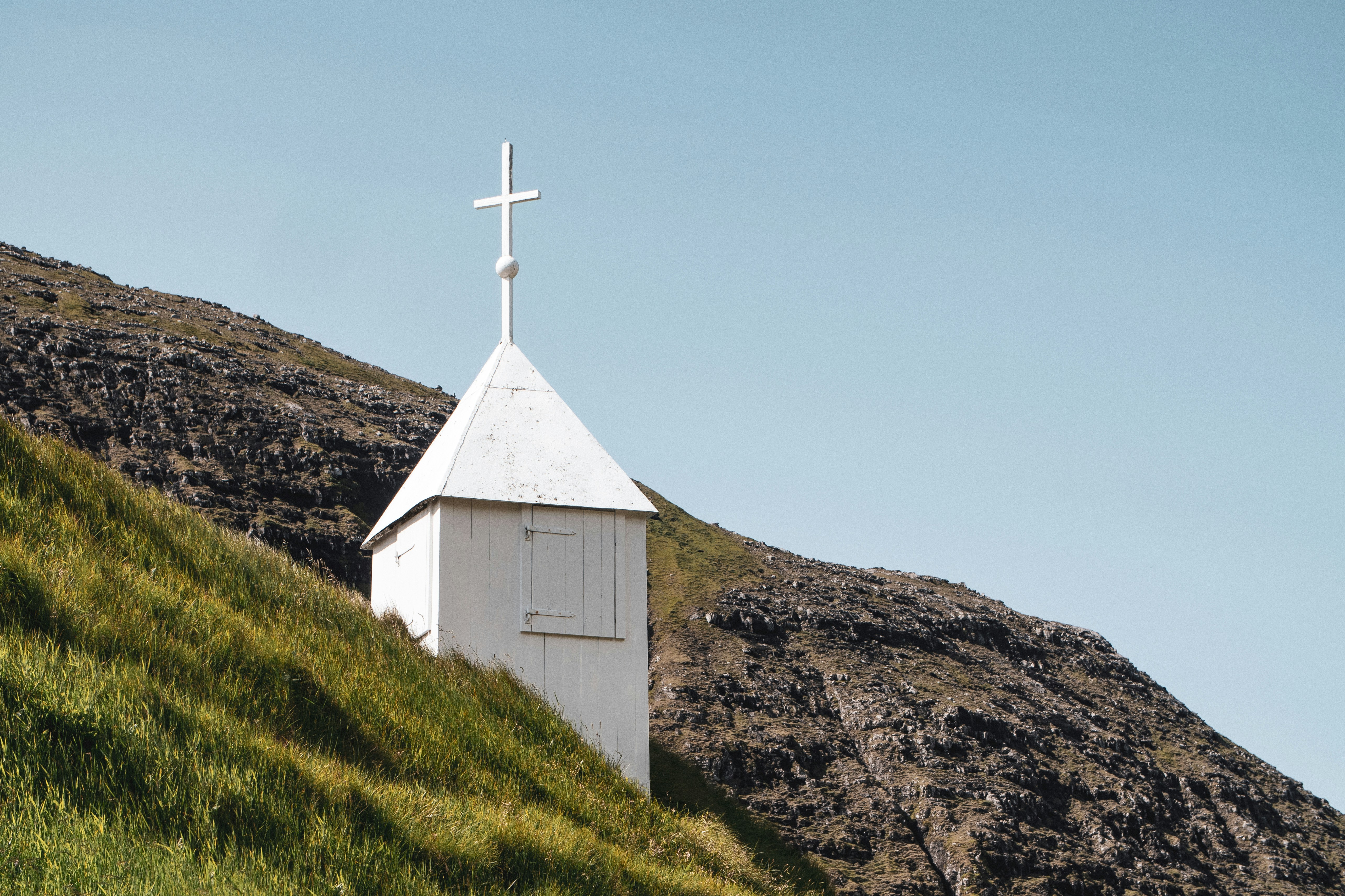 A small white church perched on a grassy slope, surrounded by rugged hills under a clear blue sky.