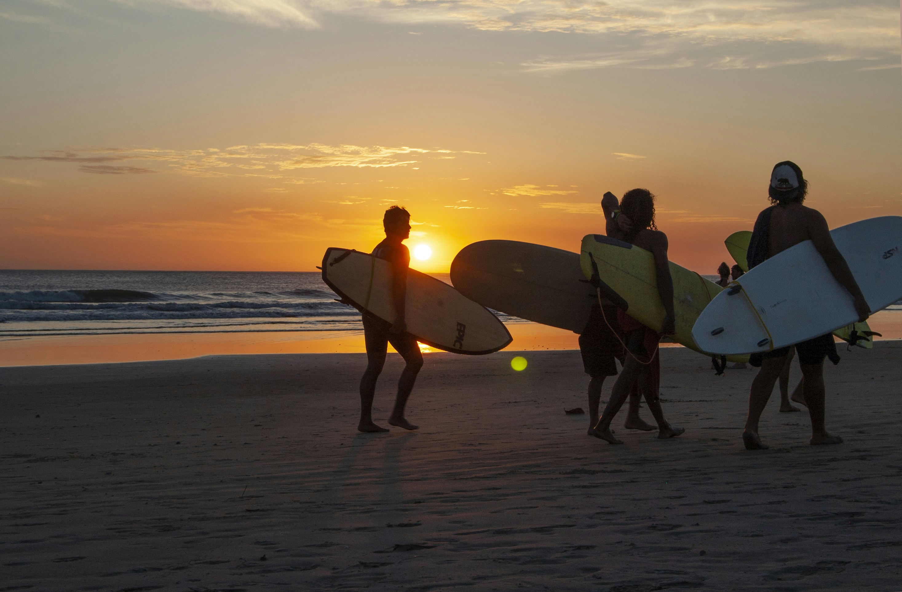 Surfers carrying boards along the shoreline as the sun sets over the ocean.