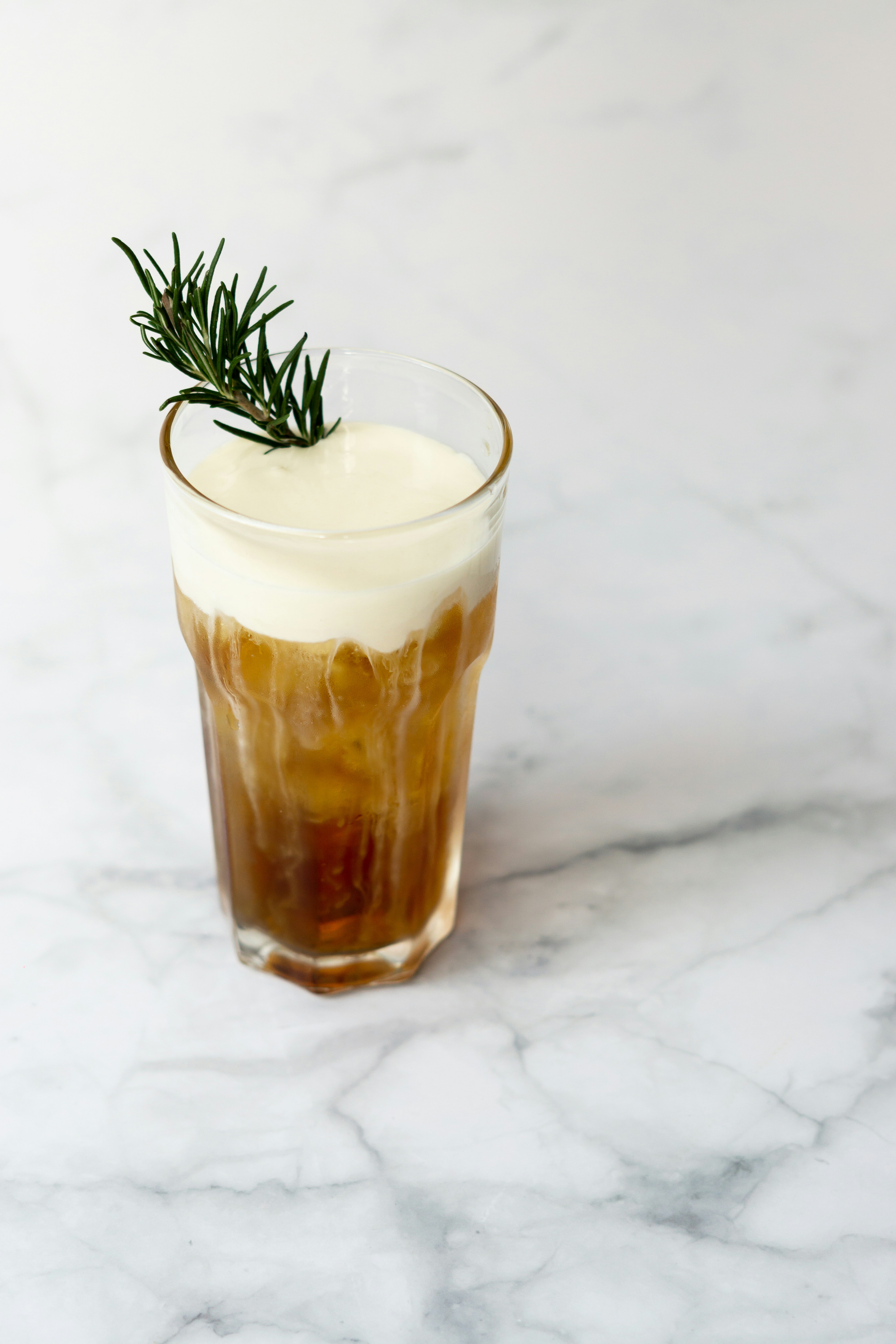 Photograph of a rosemary-garnished amber beverage in a tall glass with frothy crema on a white marble surface. The composition emphasizes the drink's garnish and the clean marble backdrop.
