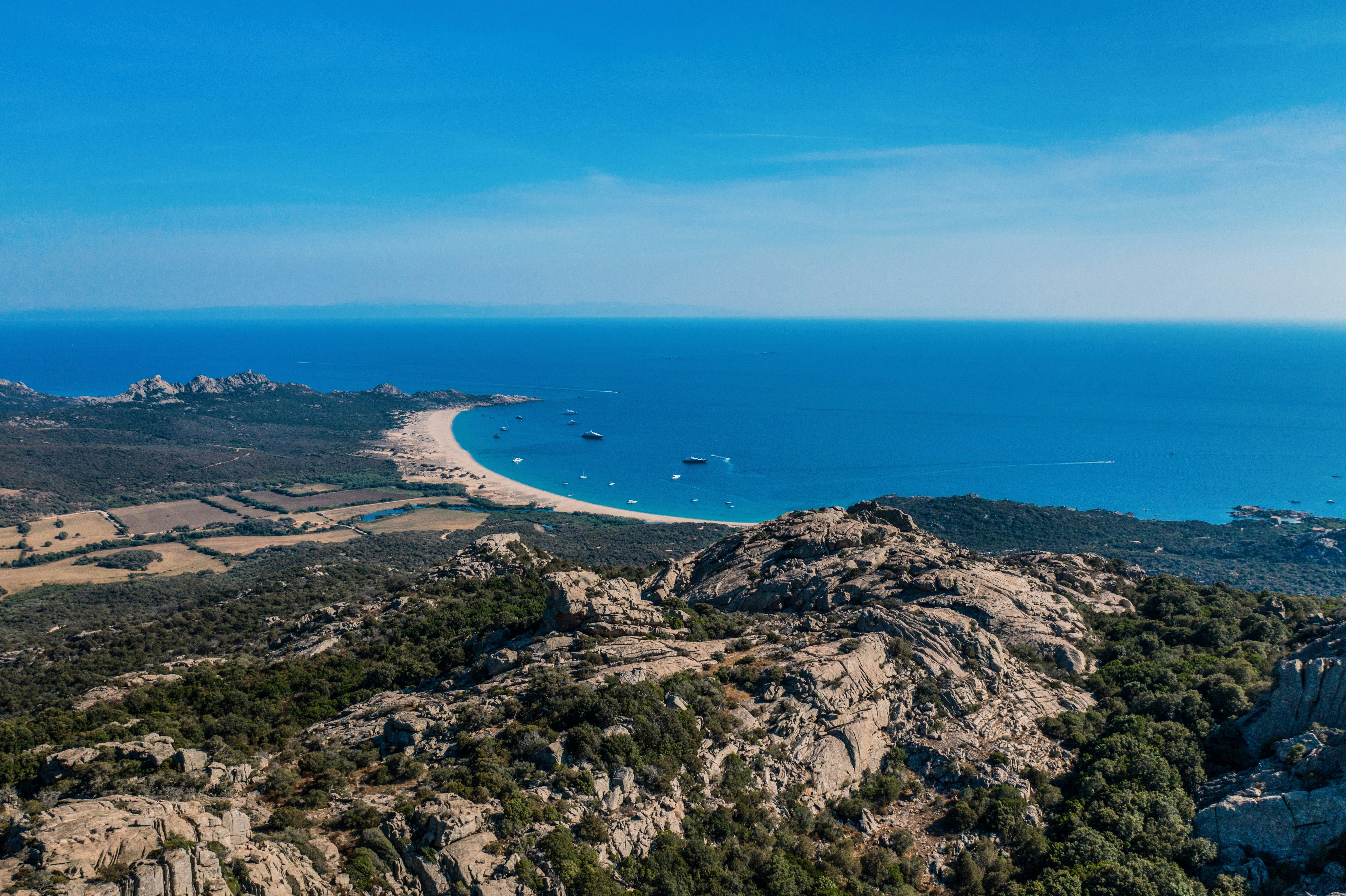Aerial view of a serene Mediterranean coastline, showcasing a sandy beach curving along the turquoise sea, framed by rocky hills and lush greenery.