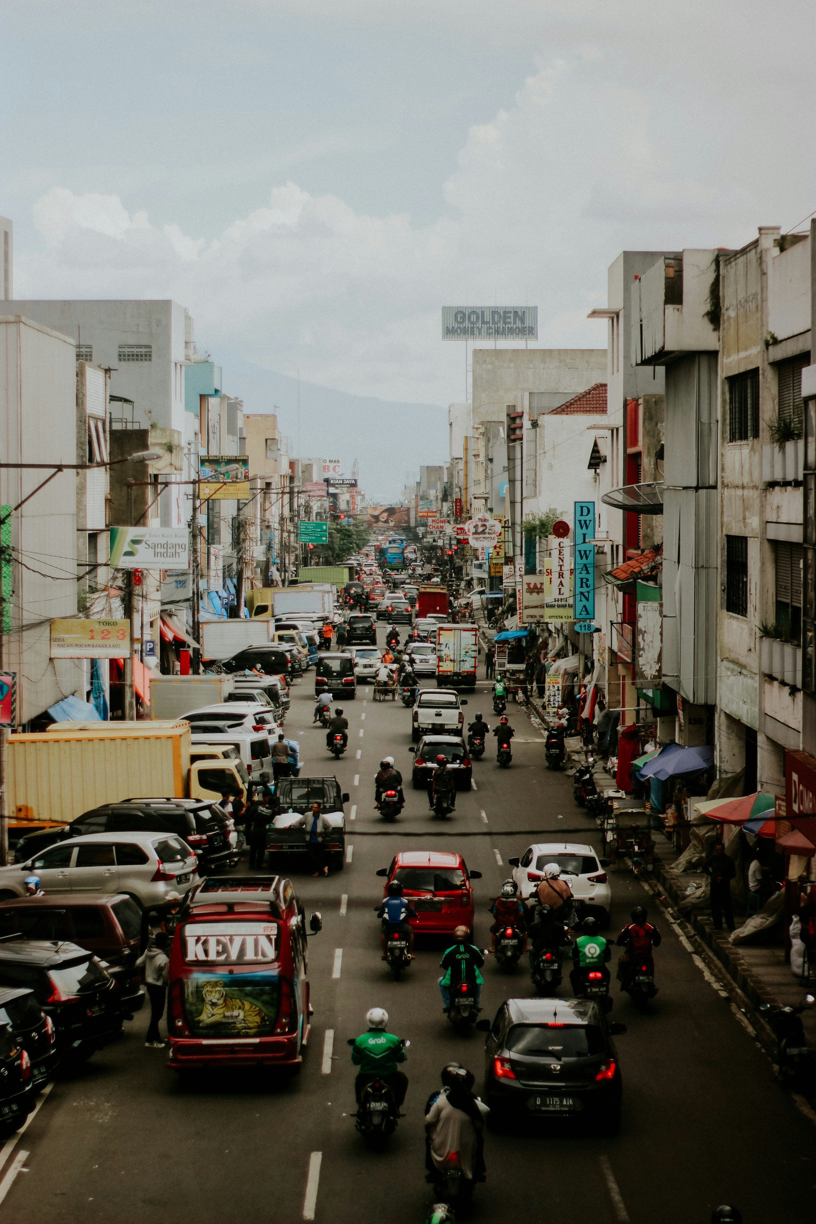 Busy street scene filled with a mix of vehicles and pedestrians in a vibrant urban setting. Signs and storefronts line the road, showcasing local culture.