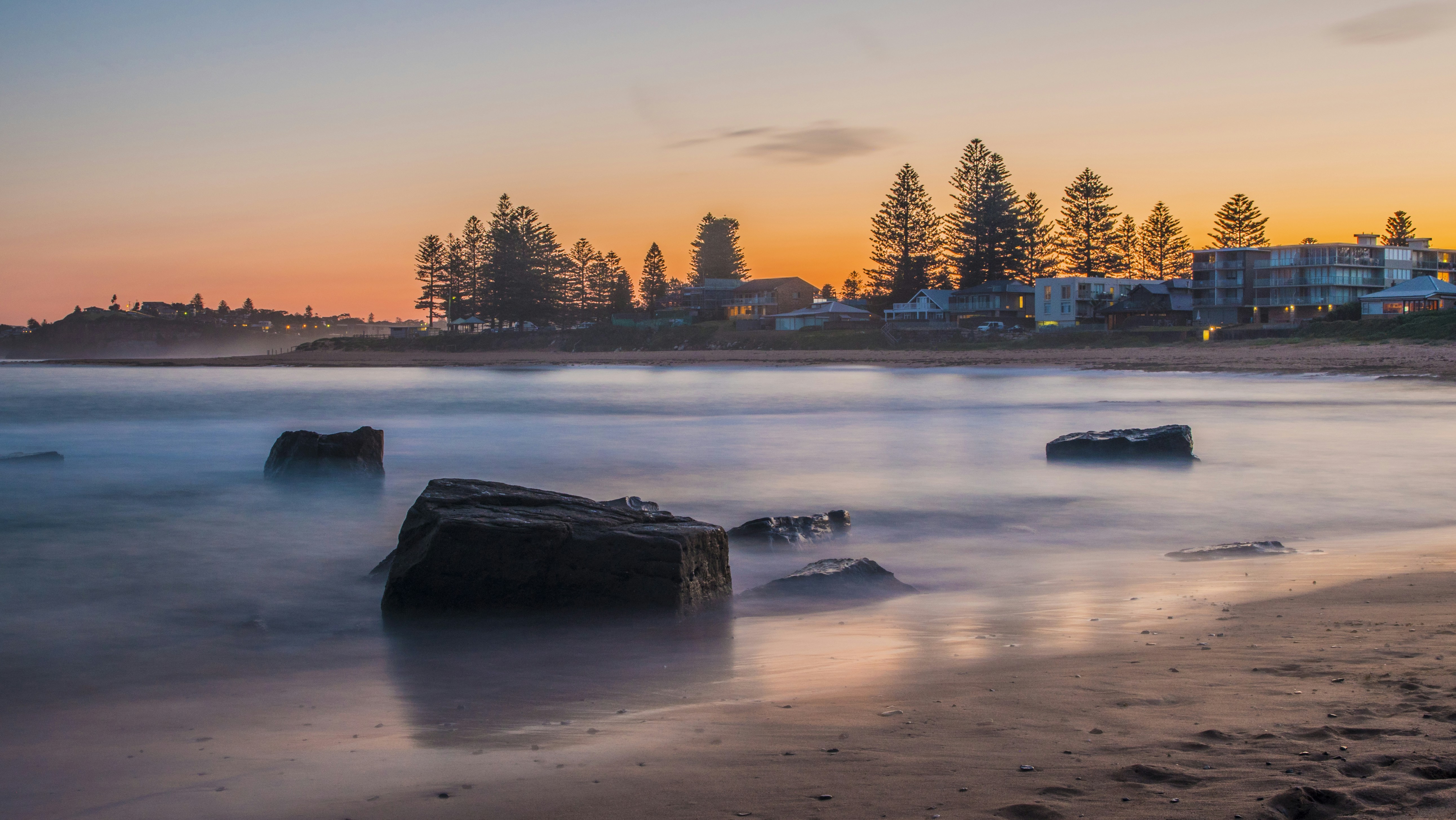 Long exposure of waves smoothing over rocks at Mona Vale Beach during sunset, with silhouettes of trees and buildings on the horizon.