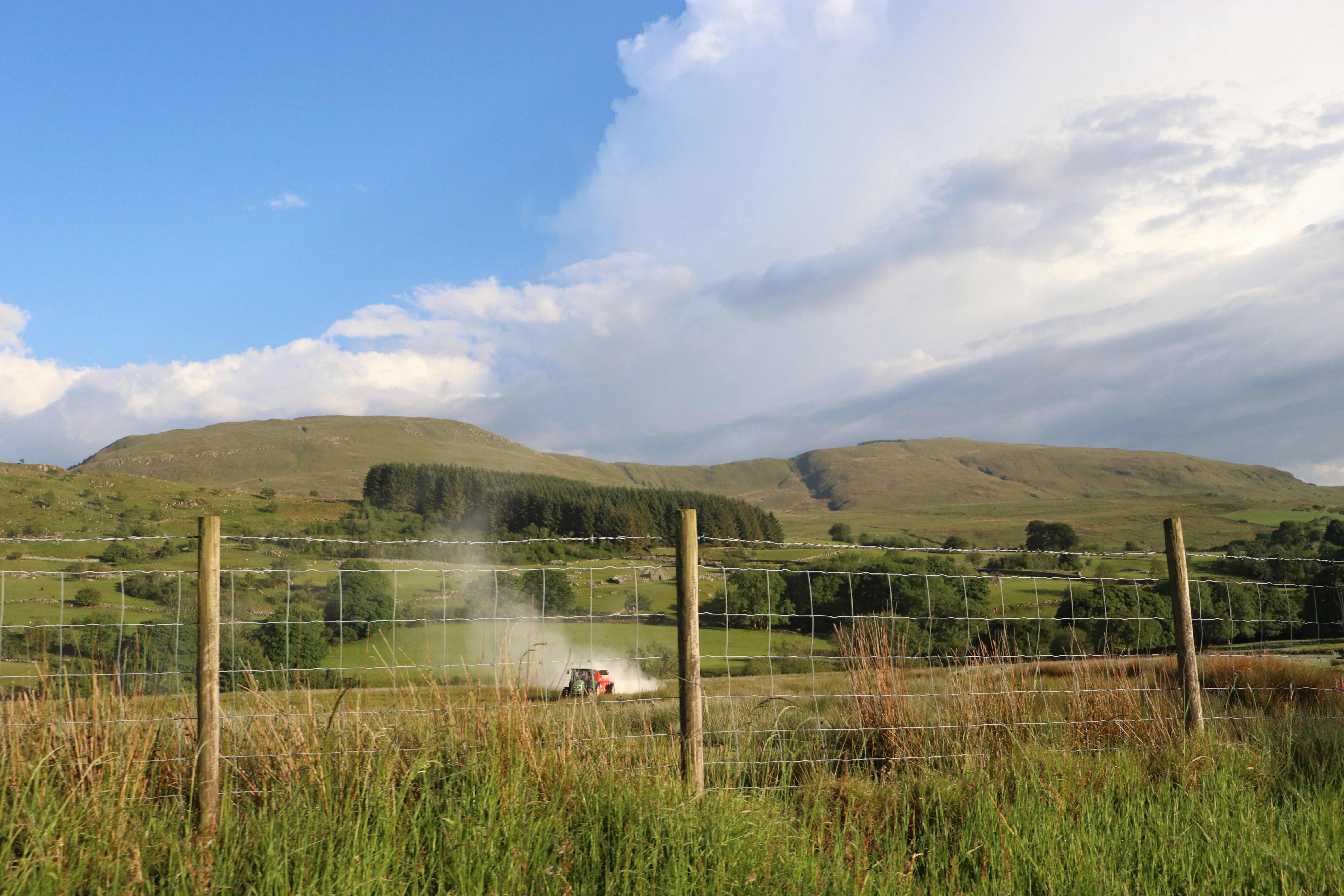Tractor kicking up dust on a rural road, framed by lush green hills and a dramatic sky. The scene captures the essence of pastoral life in the Highlands.