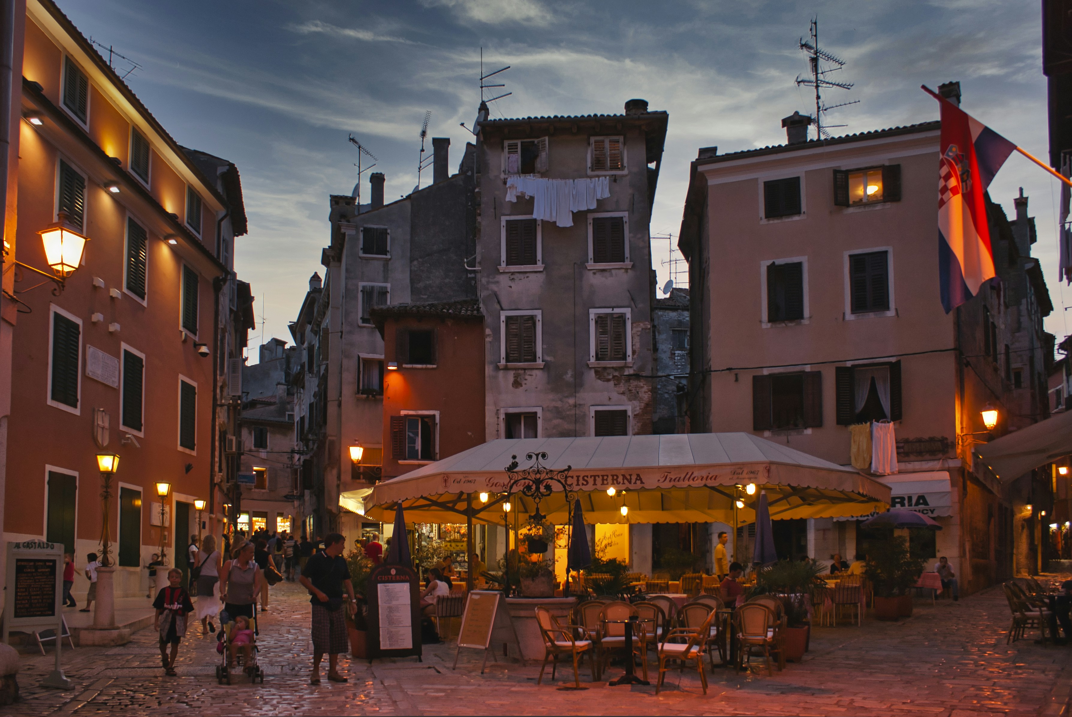 People stroll past a warmly lit trattoria in a cobblestone square under a twilight sky.