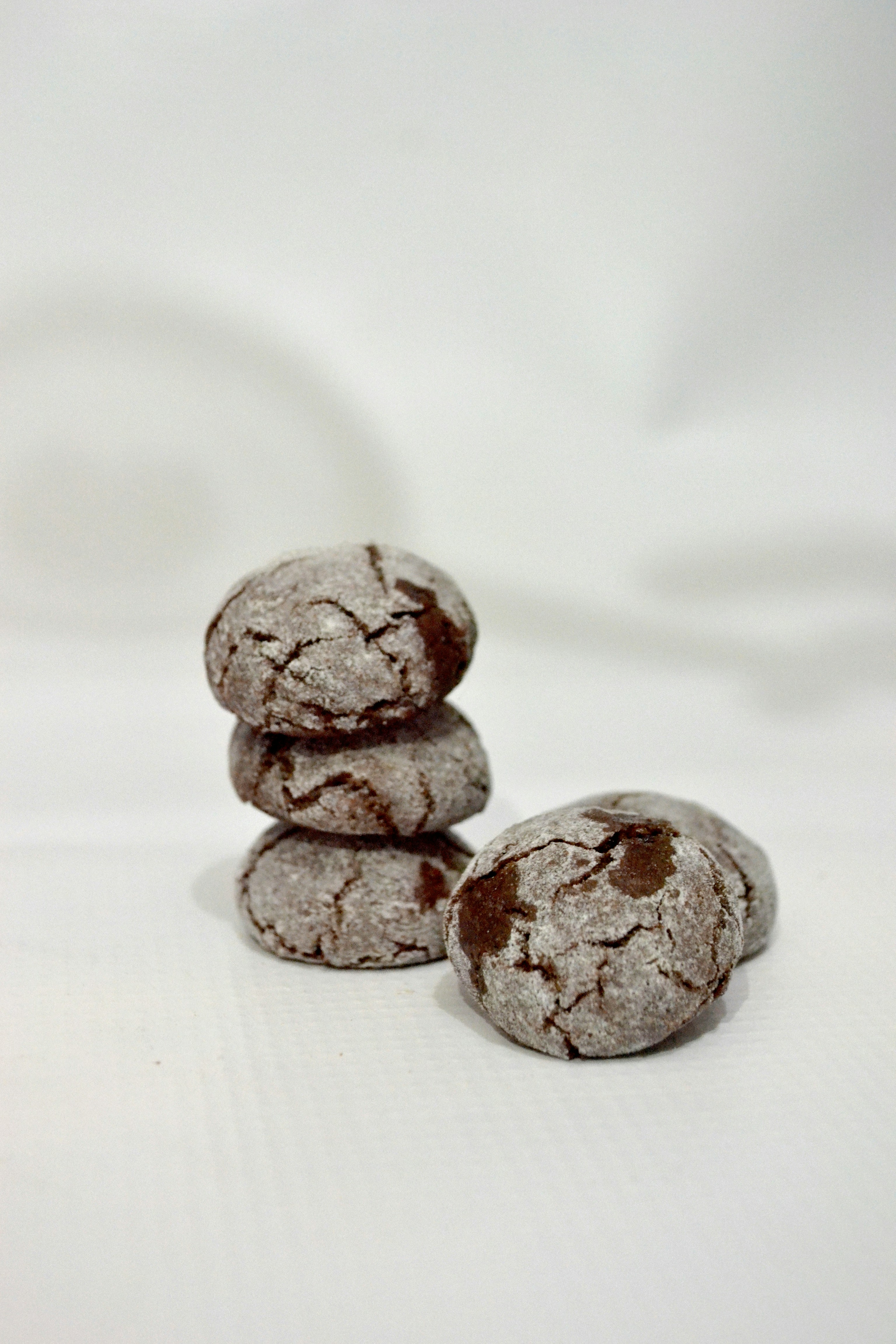 Four chocolate crinkle cookies stacked with a dusting of powdered sugar, set against a soft white background.