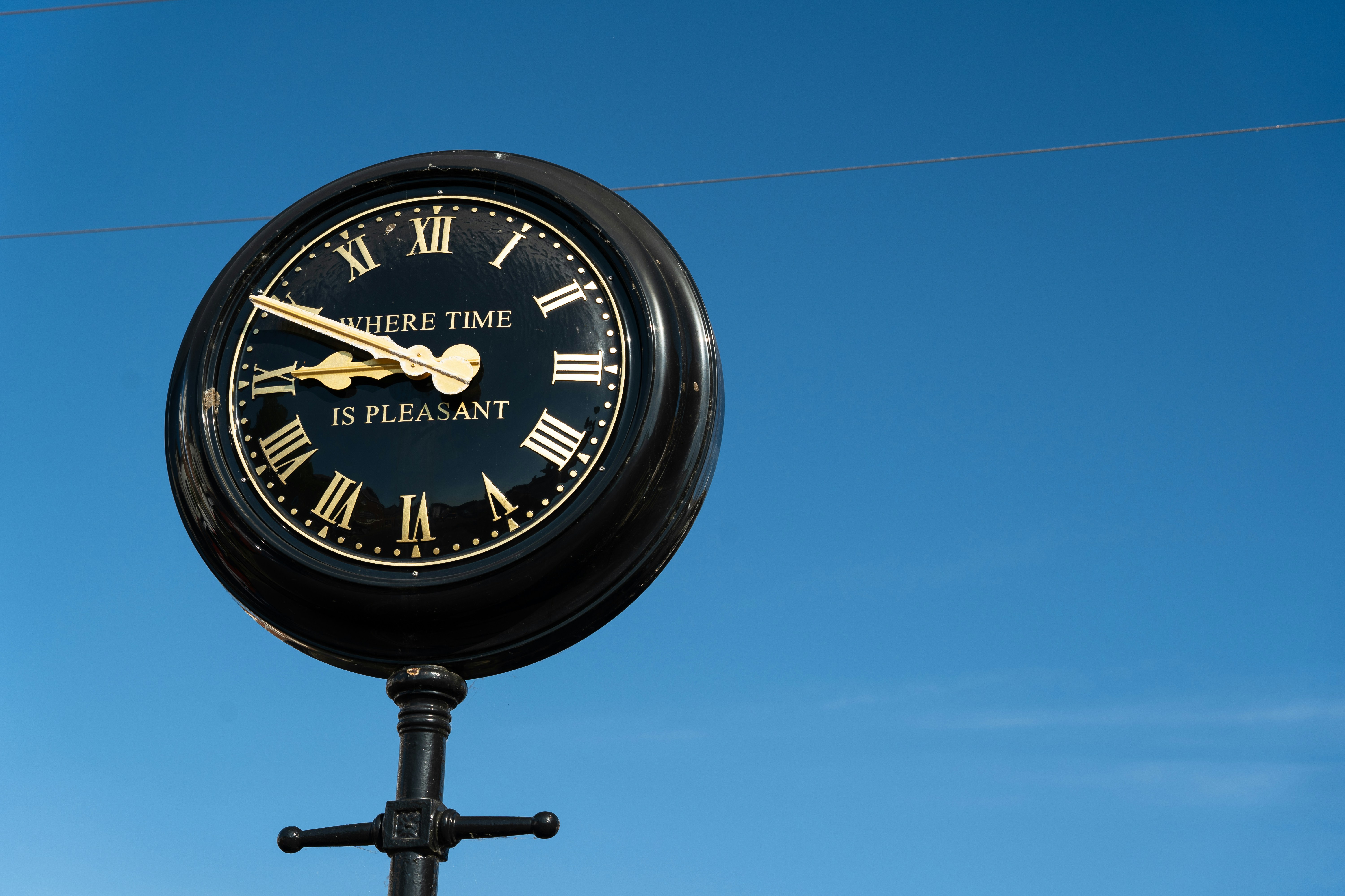 Elegant street clock displaying Roman numerals against a clear blue sky, emphasizing the phrase 'WHERE TIME IS PLEASANT.'