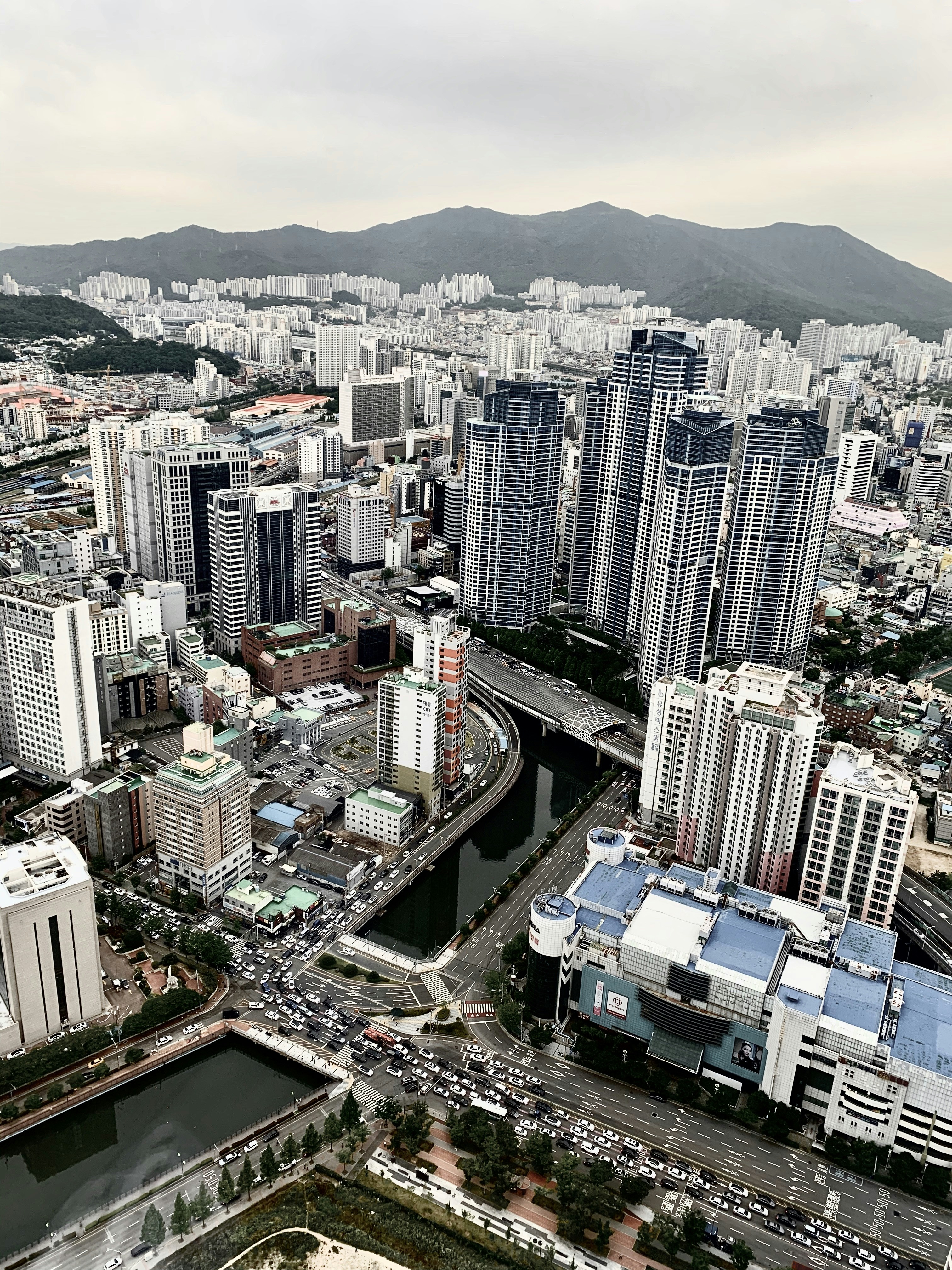 Aerial view of a bustling cityscape showcasing a blend of modern skyscrapers and winding roads alongside a river. The backdrop features distant mountains under a cloudy sky.