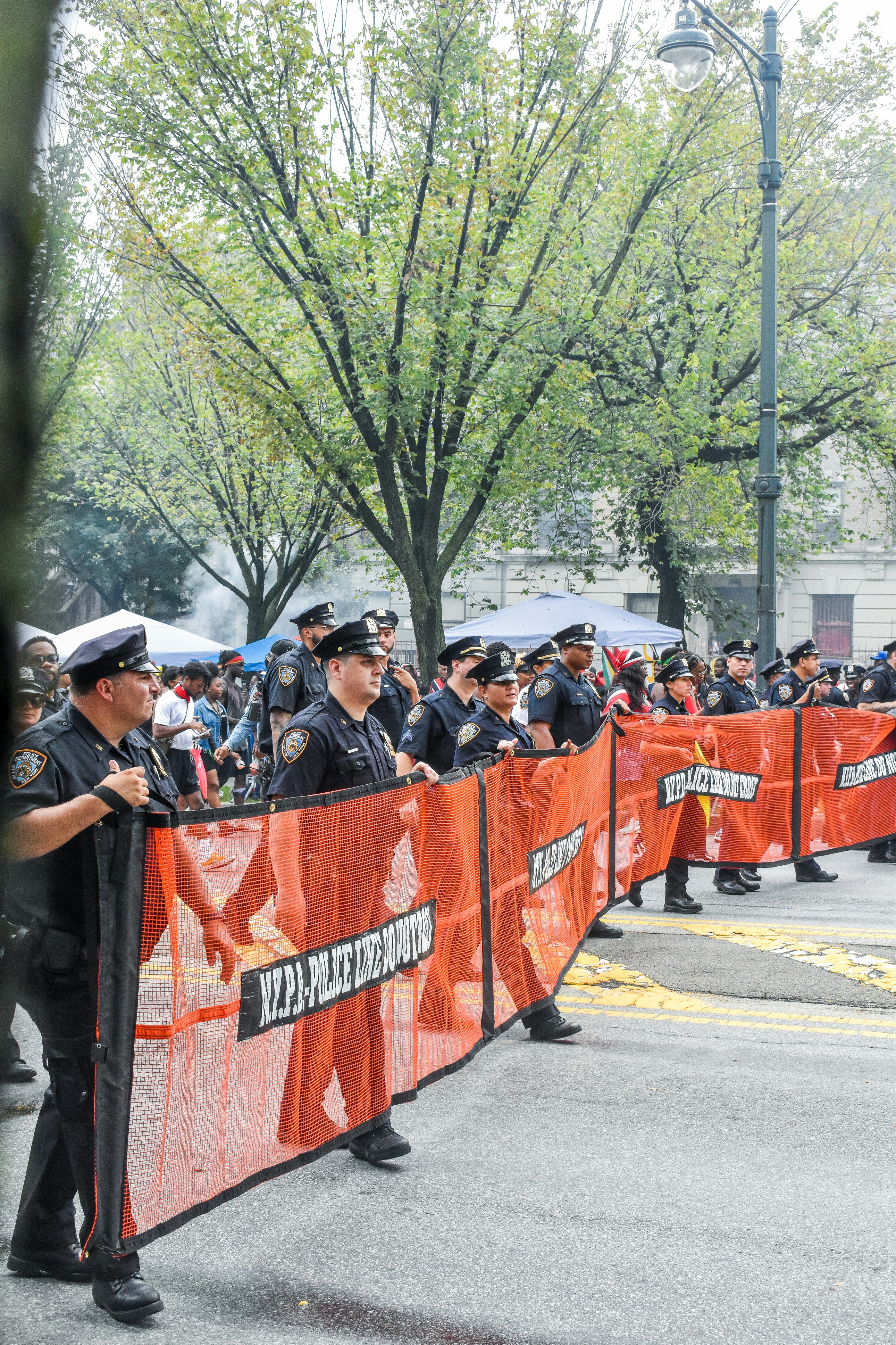Police officers standing resolutely behind a barricade during a public event, showcasing their role in maintaining order. The scene is set against a backdrop of trees and tents.