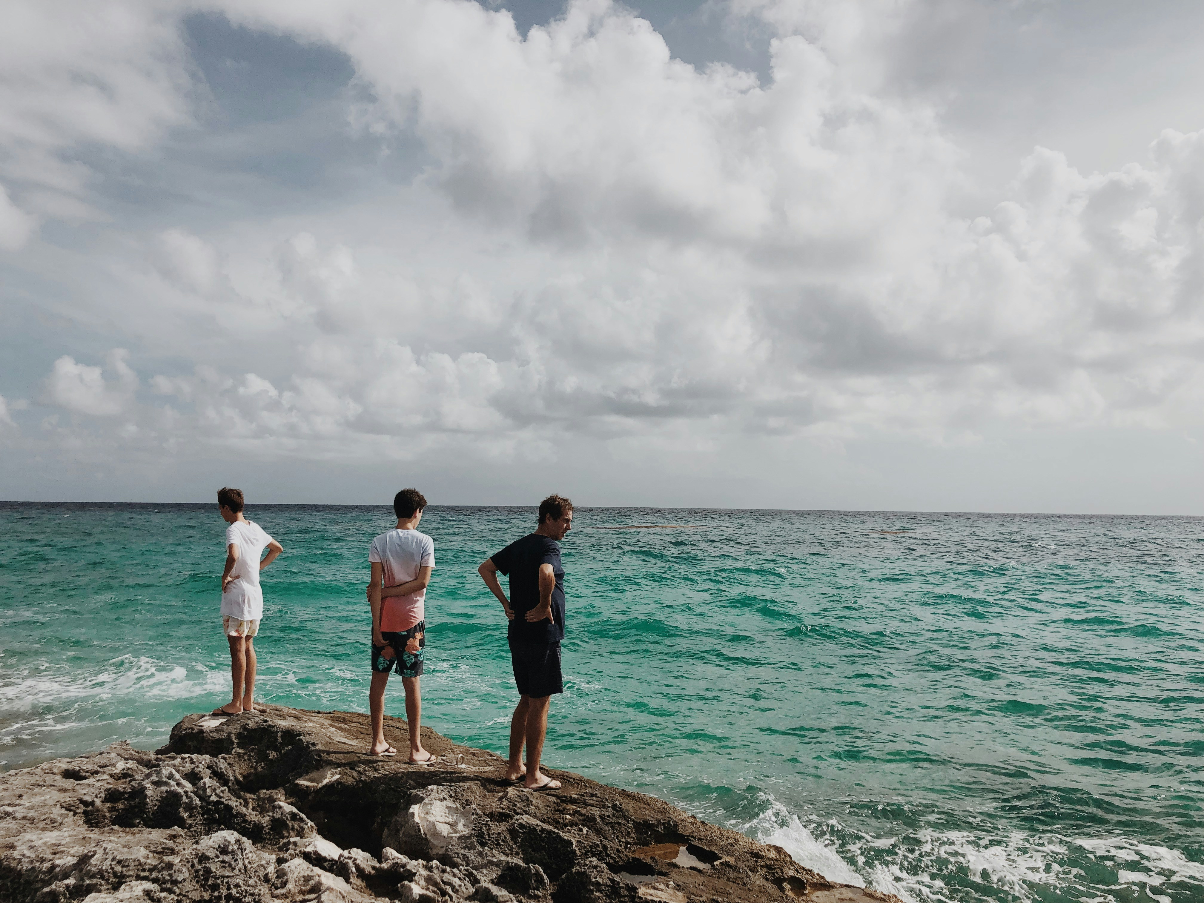 Three people stand on a rocky shore gazing out at the turquoise sea under a cloudy sky.