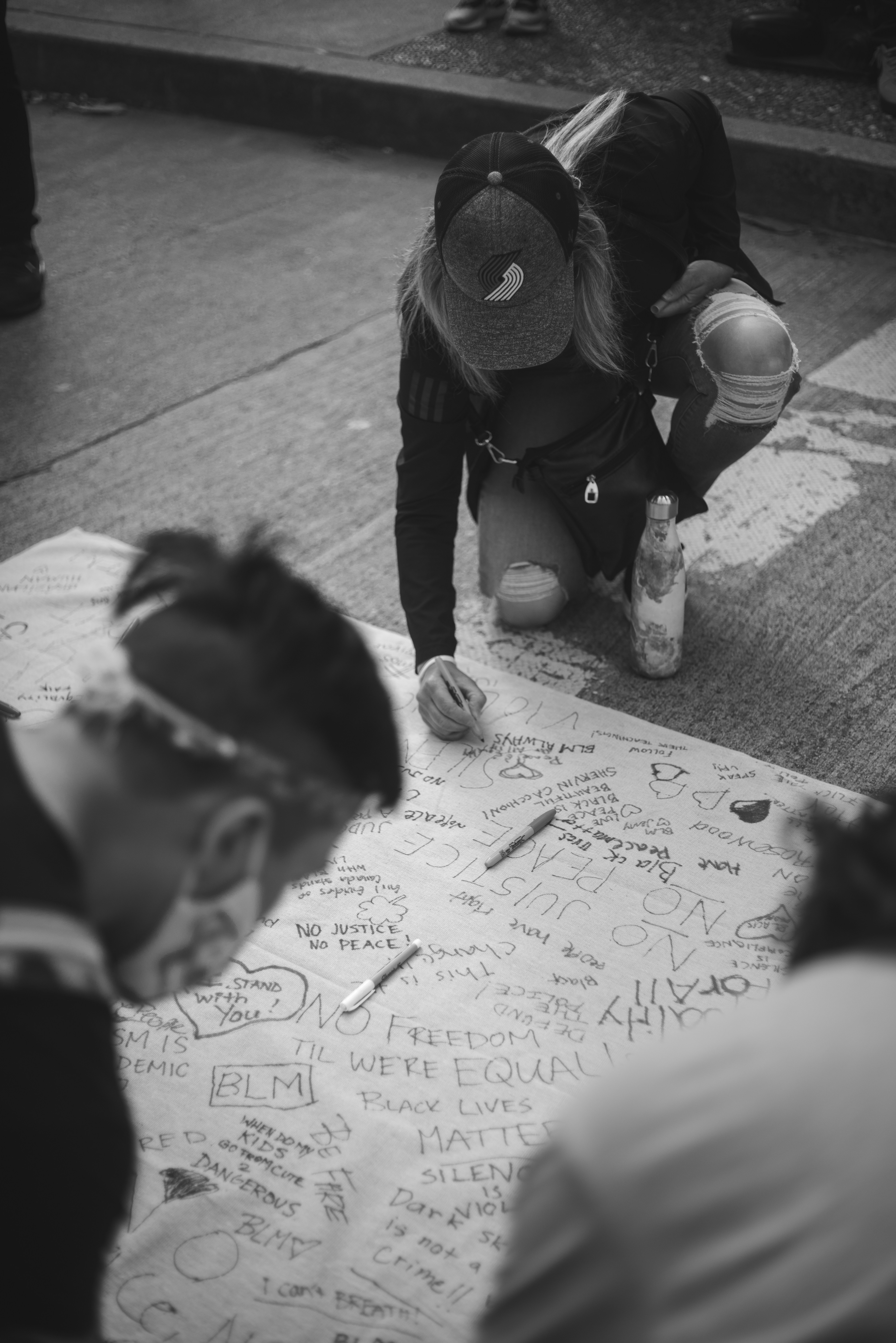 Participants writing messages on a large sheet during a protest, emphasizing social justice themes. The scene captures the spirit of activism and community engagement.