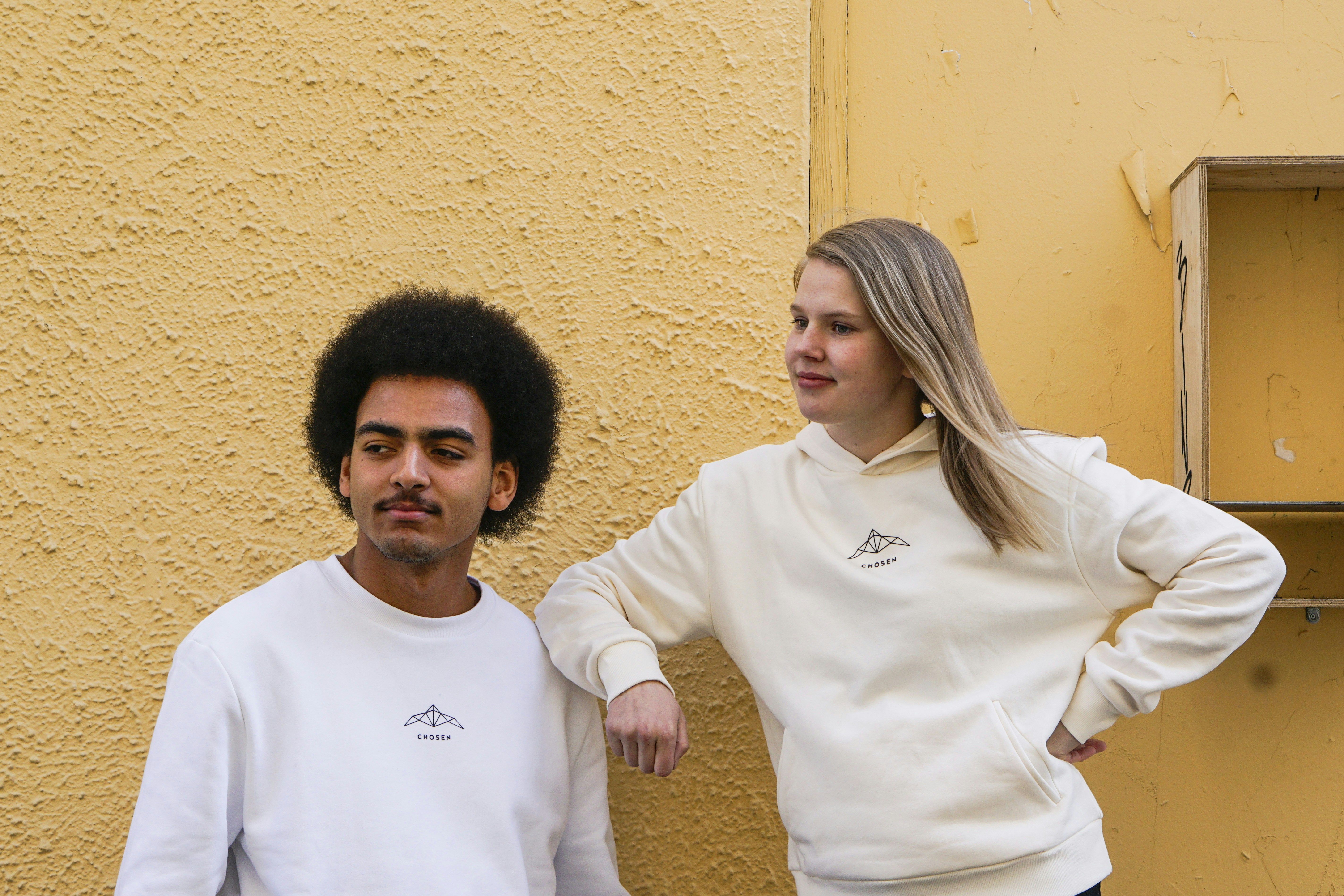 Two people in casual outfits stand against a yellow textured wall.