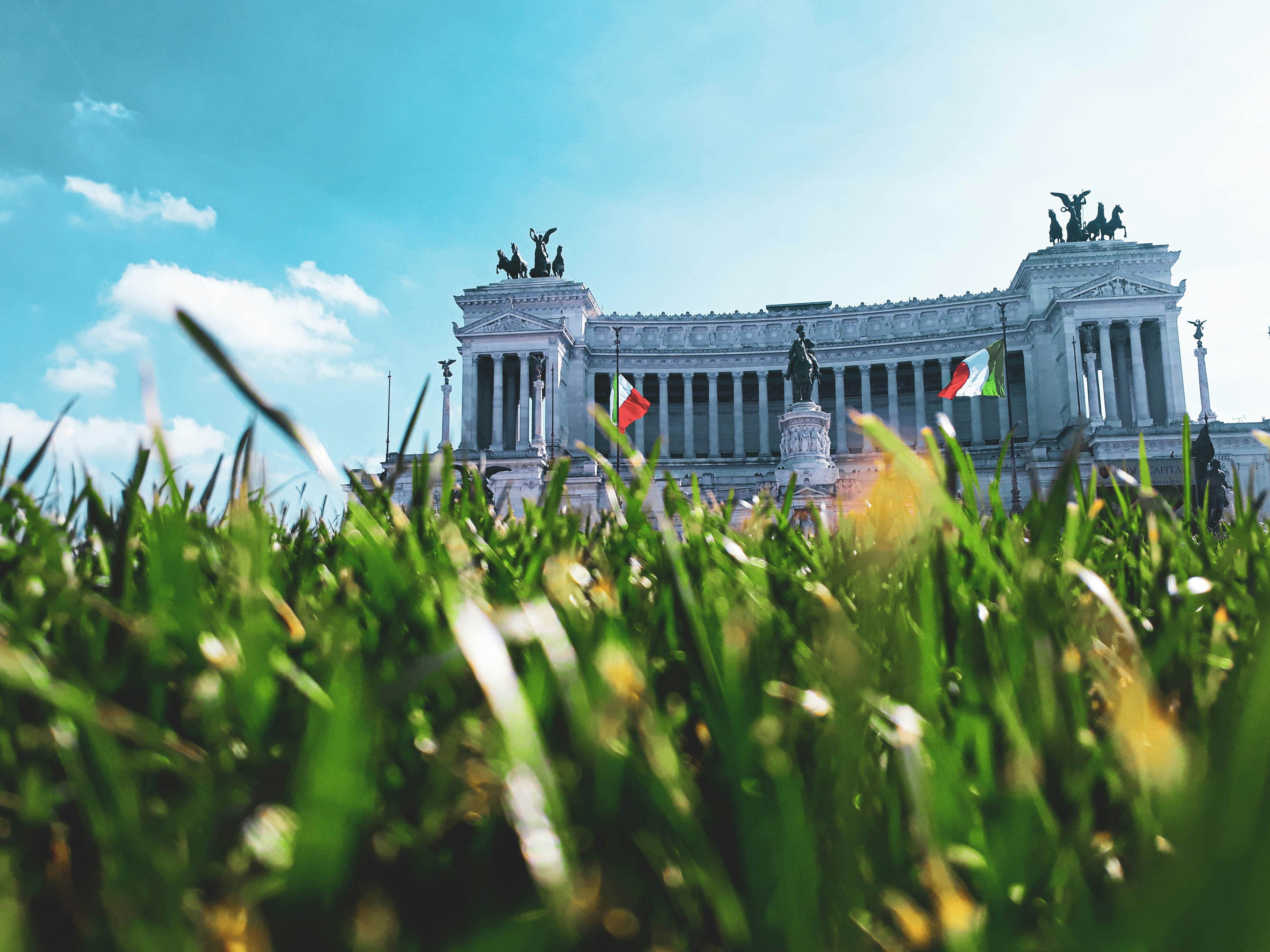 Low-angle photograph of Rome's Vittoriano monument with statues and Italian flags, framed by foreground grass against a bright blue sky.