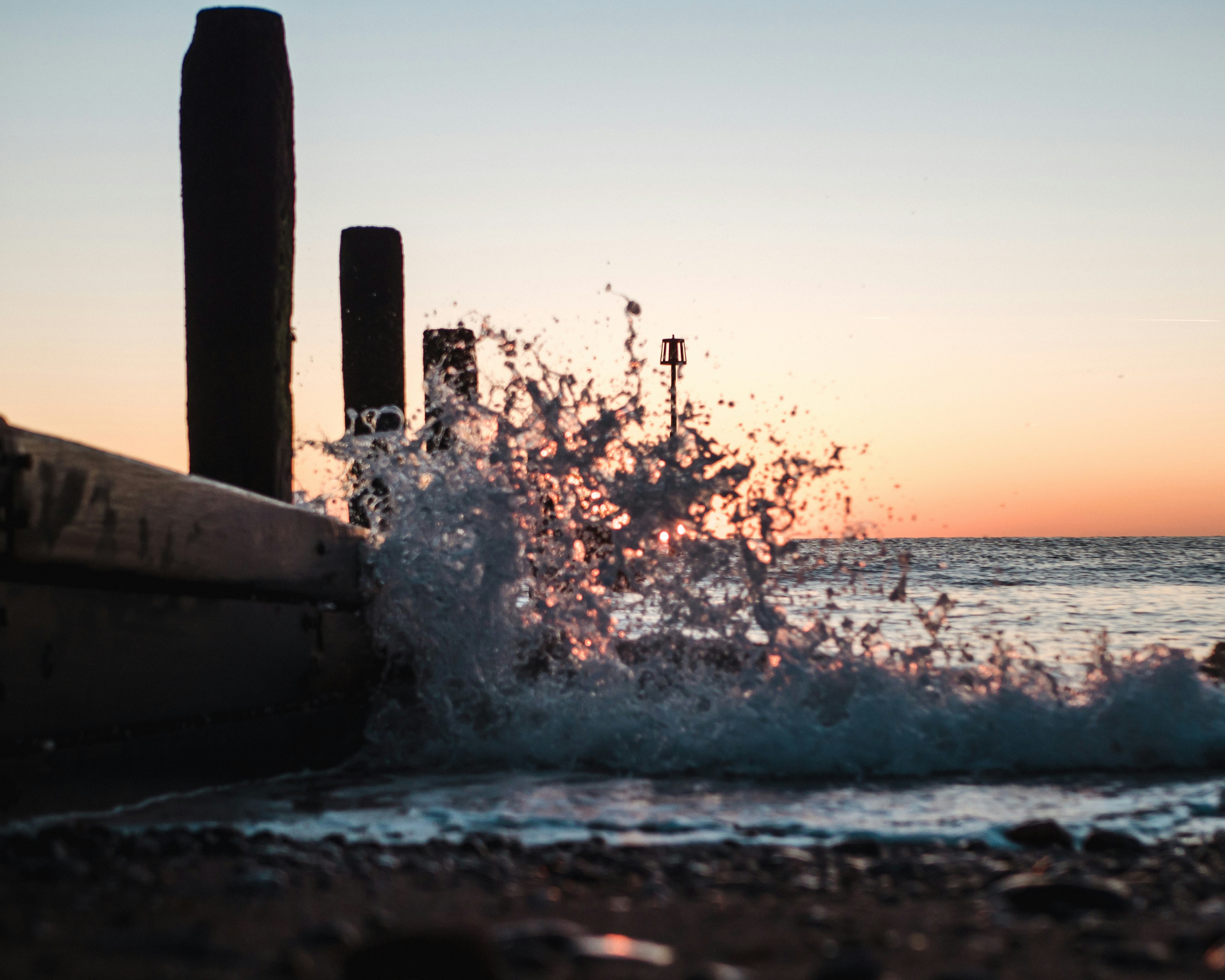 Waves crashing against wooden posts on a beach at sunrise, with vibrant hues in the sky.