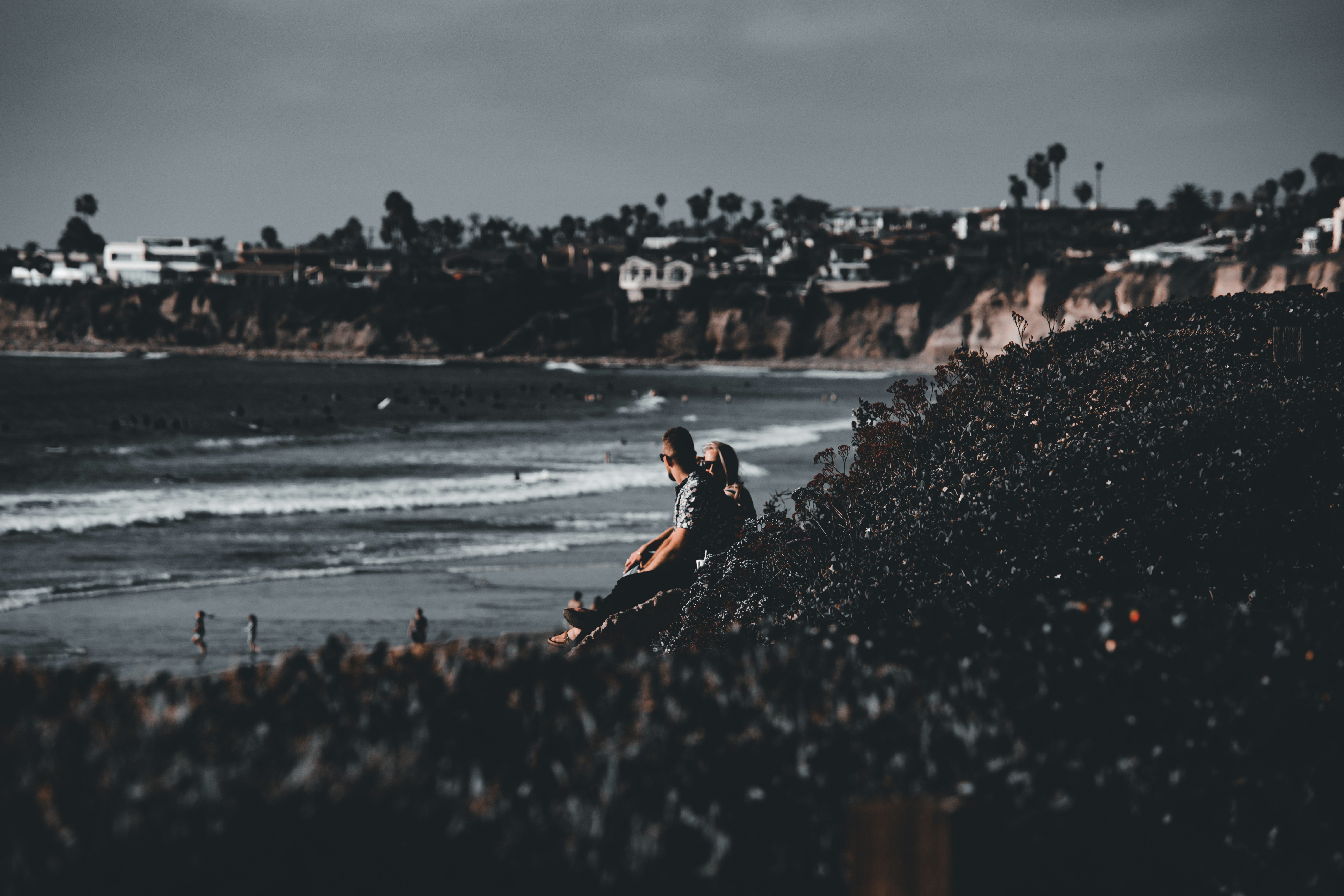 Couple sitting on a cliff overlooking the Pacific Ocean in Southern California. | man in orange shirt and black shorts sitting on rock near body of water during daytime