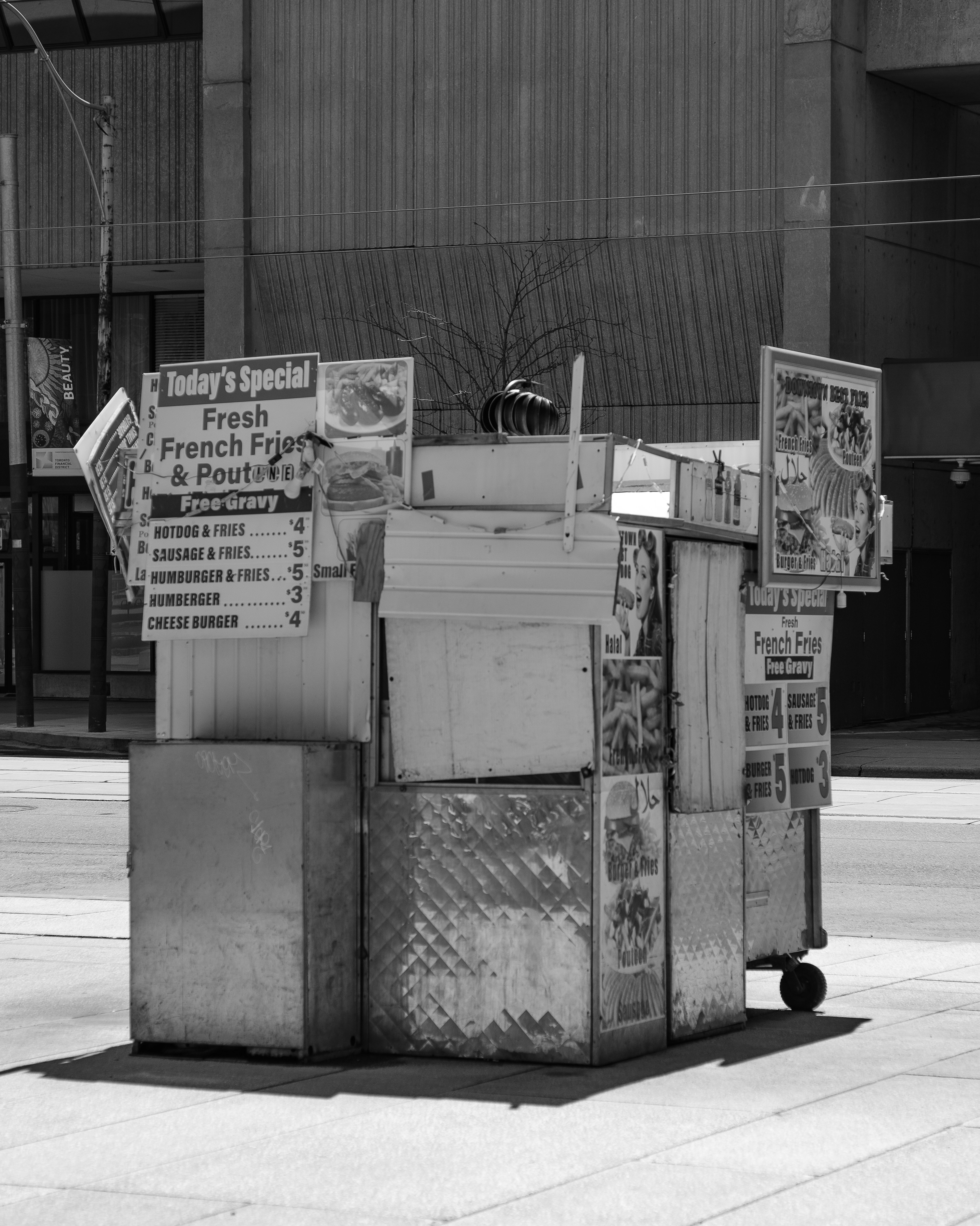 Black-and-white photograph of a street food cart with menu boards advertising fries and poutine, parked on a sidewalk with an urban backdrop.