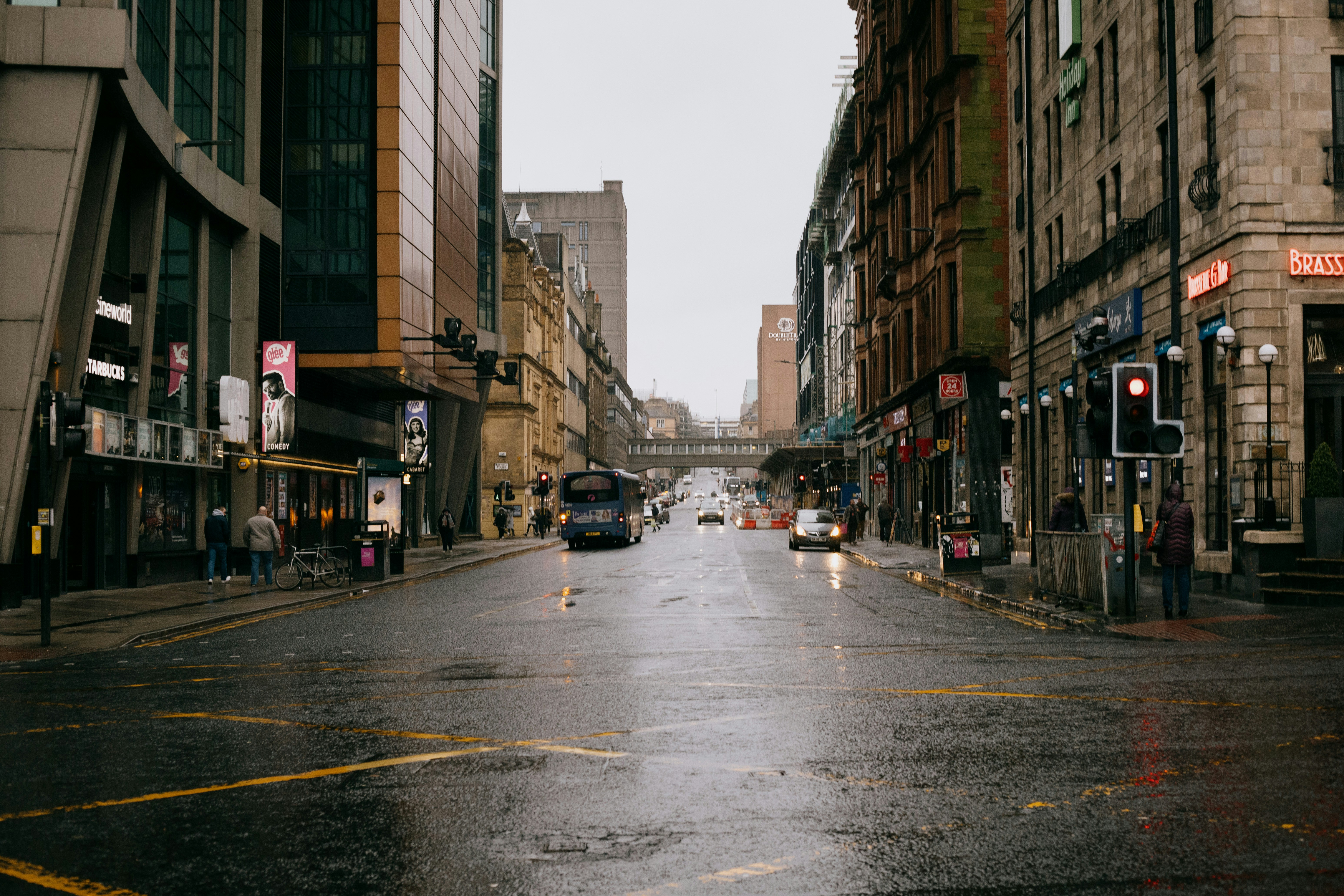 Rain-soaked city street with cars and pedestrians in Glasgow, flanked by tall buildings under a grey sky.