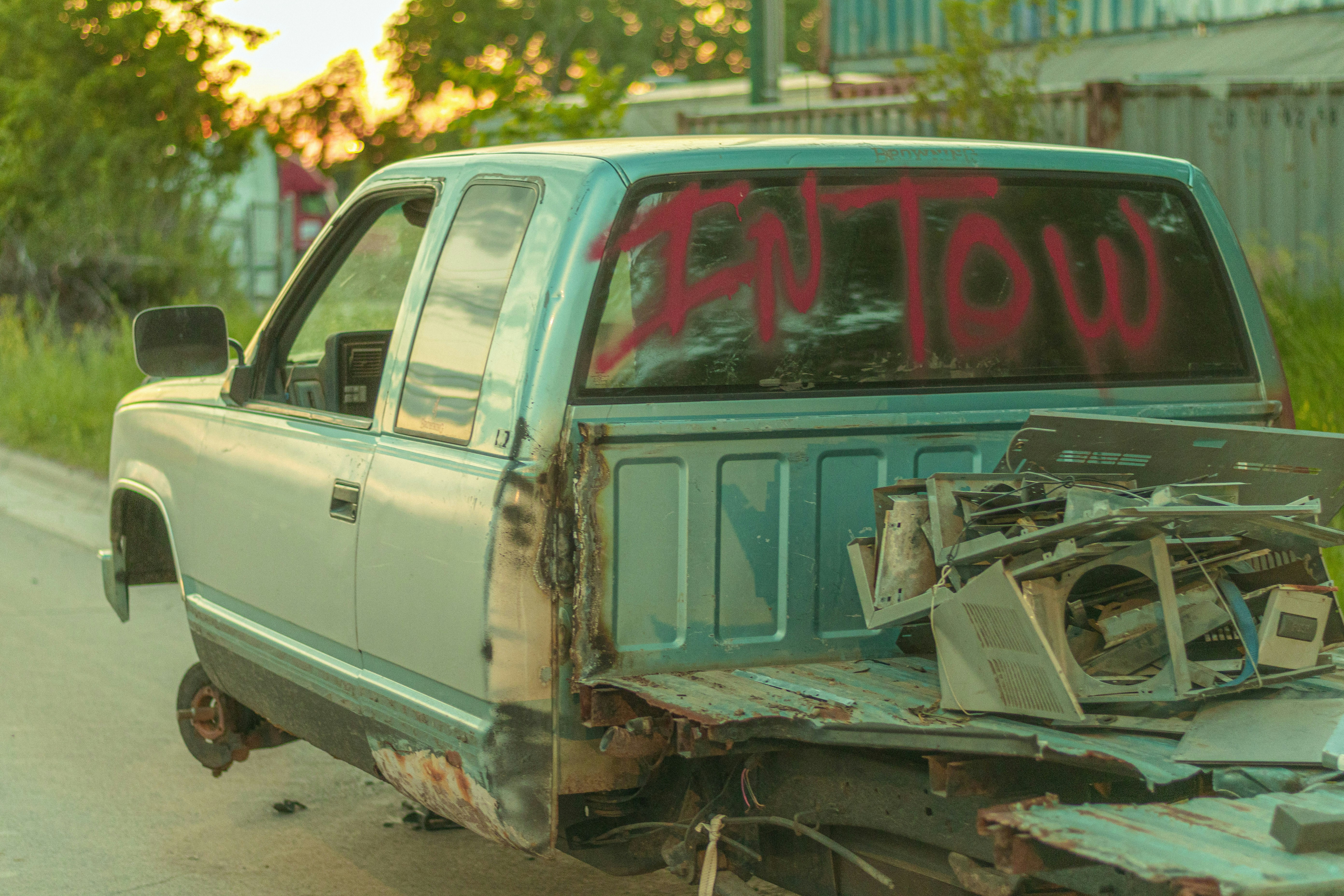 Old pickup truck with 'In Tow' spray-painted on the rear window, parked on a quiet street at sunset.