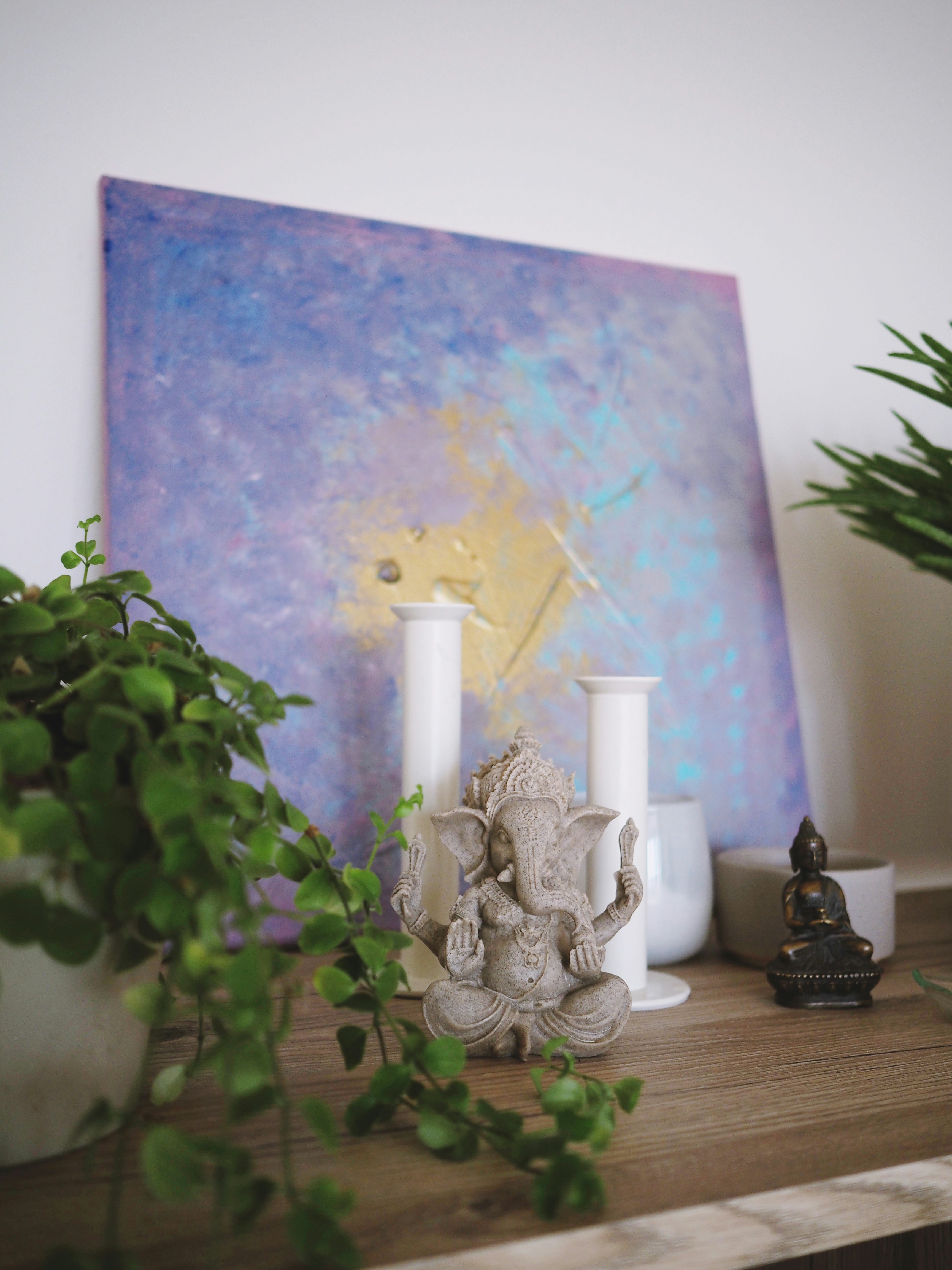 A serene altar featuring a Ganesh statue surrounded by candles and lush greenery, with an abstract painting in the background. The setup evokes tranquility and spiritual reflection.