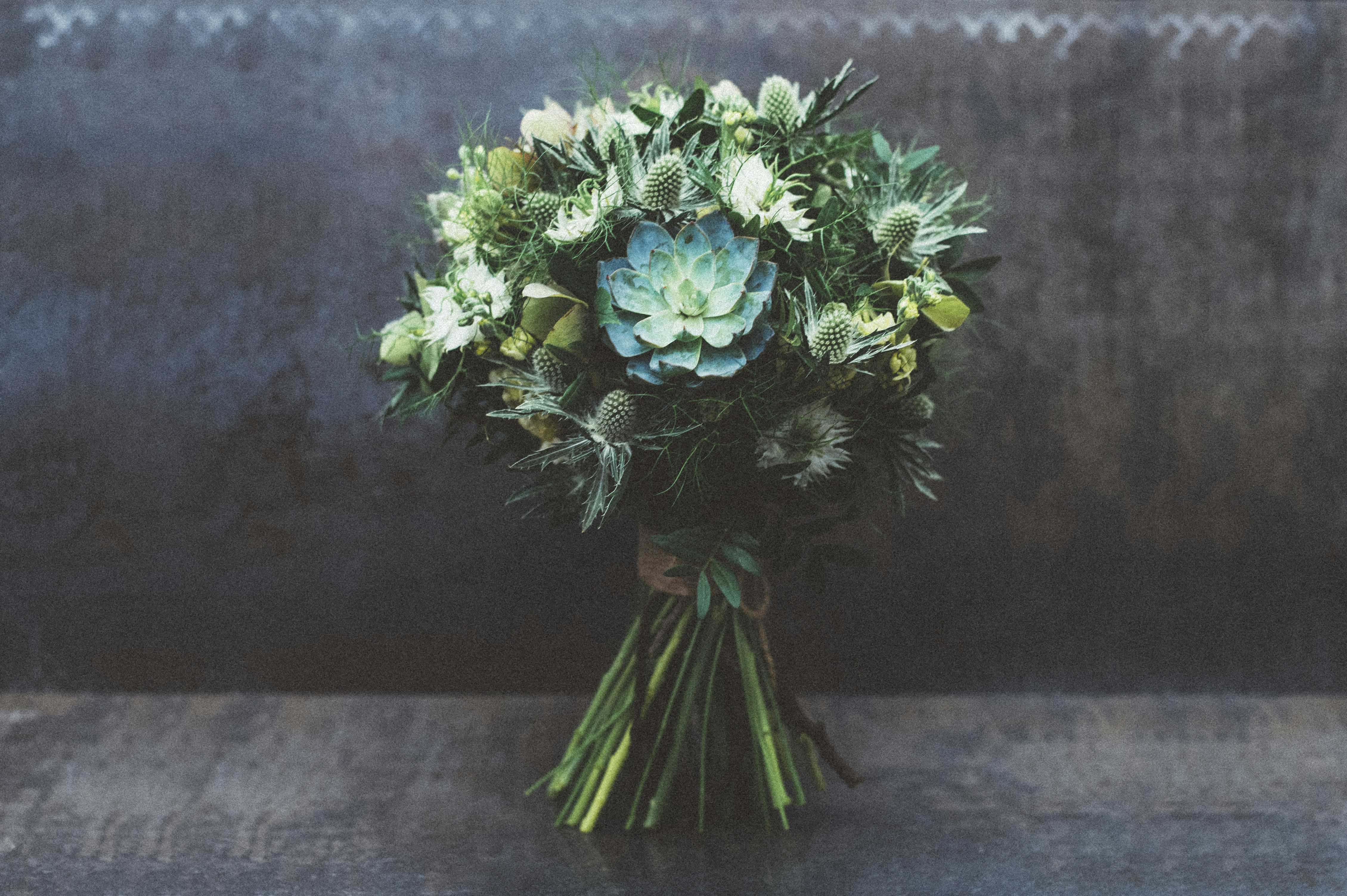 Photograph of a compact bouquet of blue-green succulents and assorted greens tied with stems, set against a dark, textured industrial backdrop.